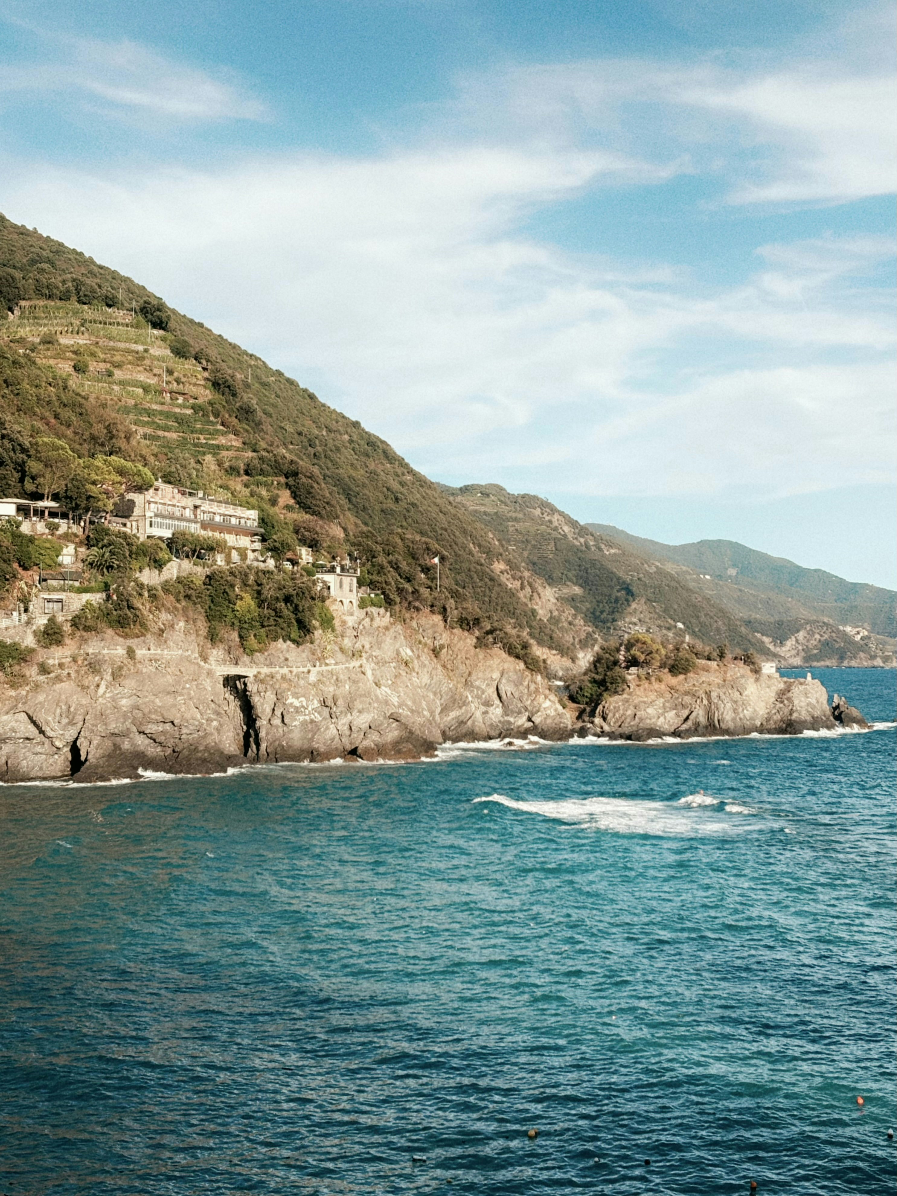 Terraced hillside coastal scene with houses perched above a rocky shoreline and calm blue sea; the composition emphasizes the cliffside architecture against a bright sky.