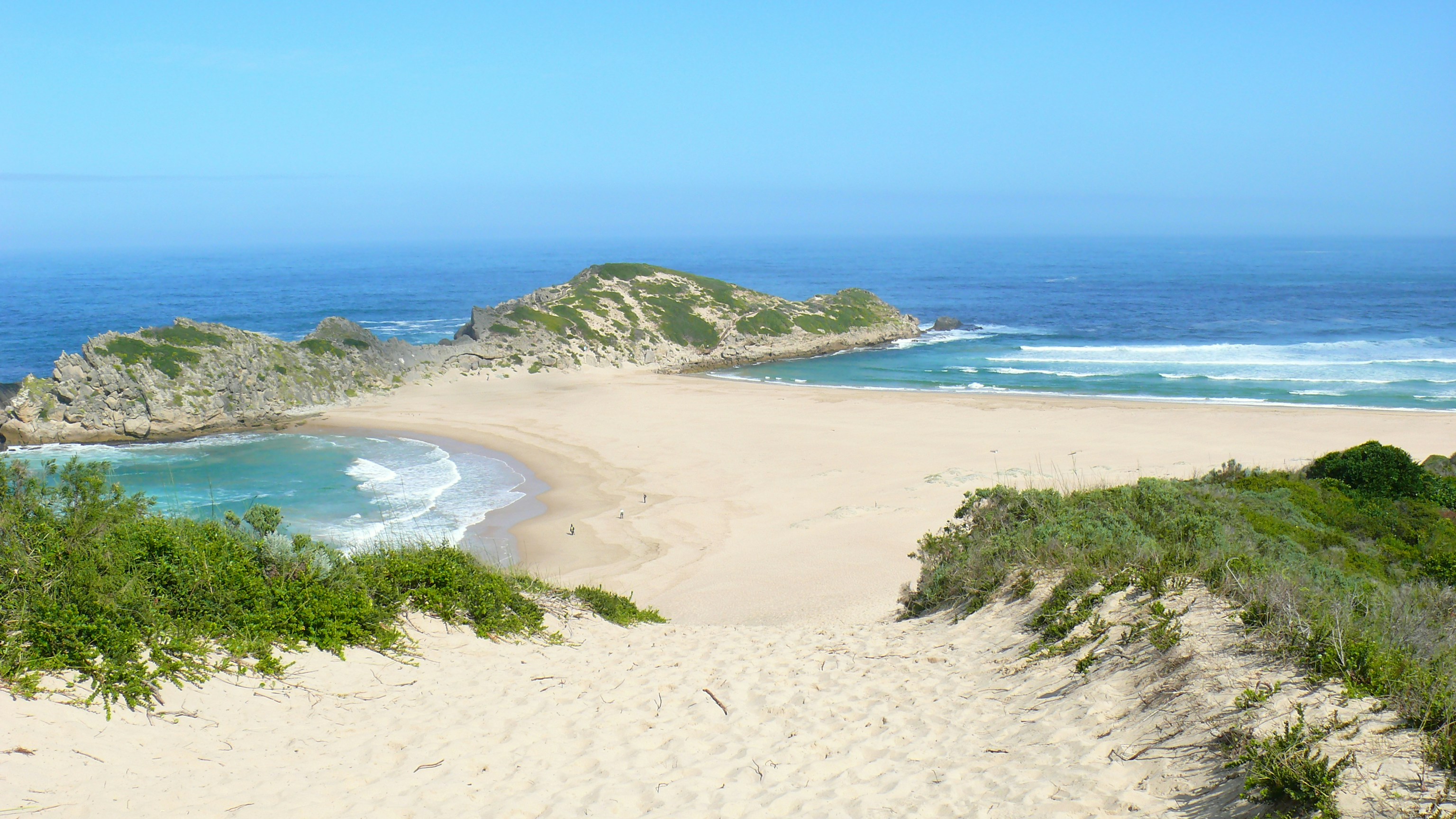 A wide, unspoiled beach stretches beneath a clear blue sky, curving around a sheltered bay where turquoise water meets pale golden sand. In the foreground, a sandy path descends through low green coastal vegetation toward the shoreline, creating a natural frame for the view. Rugged rocky headlands rise on both sides of the bay, their pale stone surfaces softened by patches of dense greenery. Gentle waves roll across the open ocean and break in white lines along the shore, while a few distant visitors emphasize the scale and peaceful isolation of this dramatic coastal landscape.