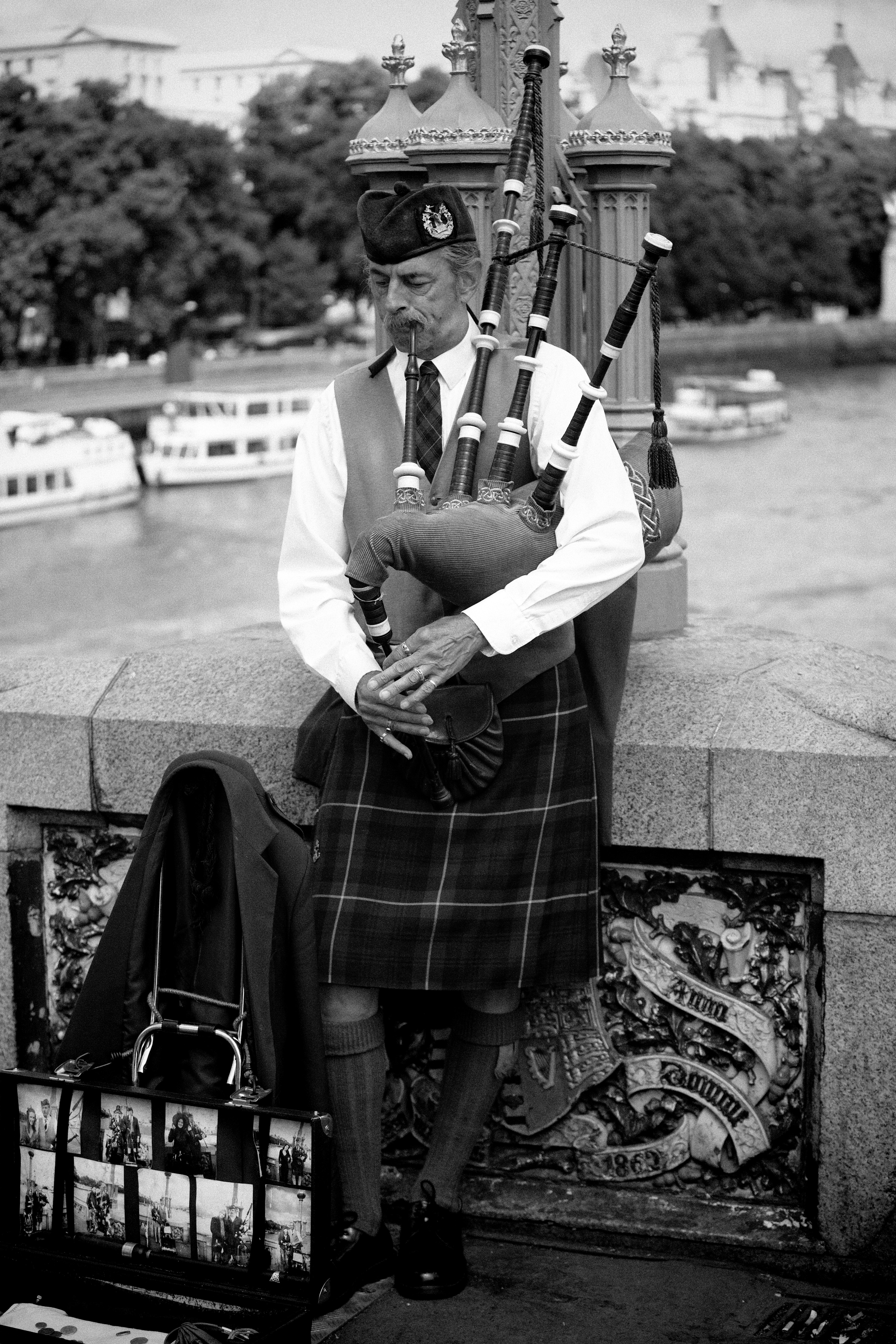Black-and-white scene of a lone bagpiper in a traditional kilt and hat, standing beside a stone railing near the river; a carry-case with photographs rests at his feet, suggesting a street performance in a historic city setting.