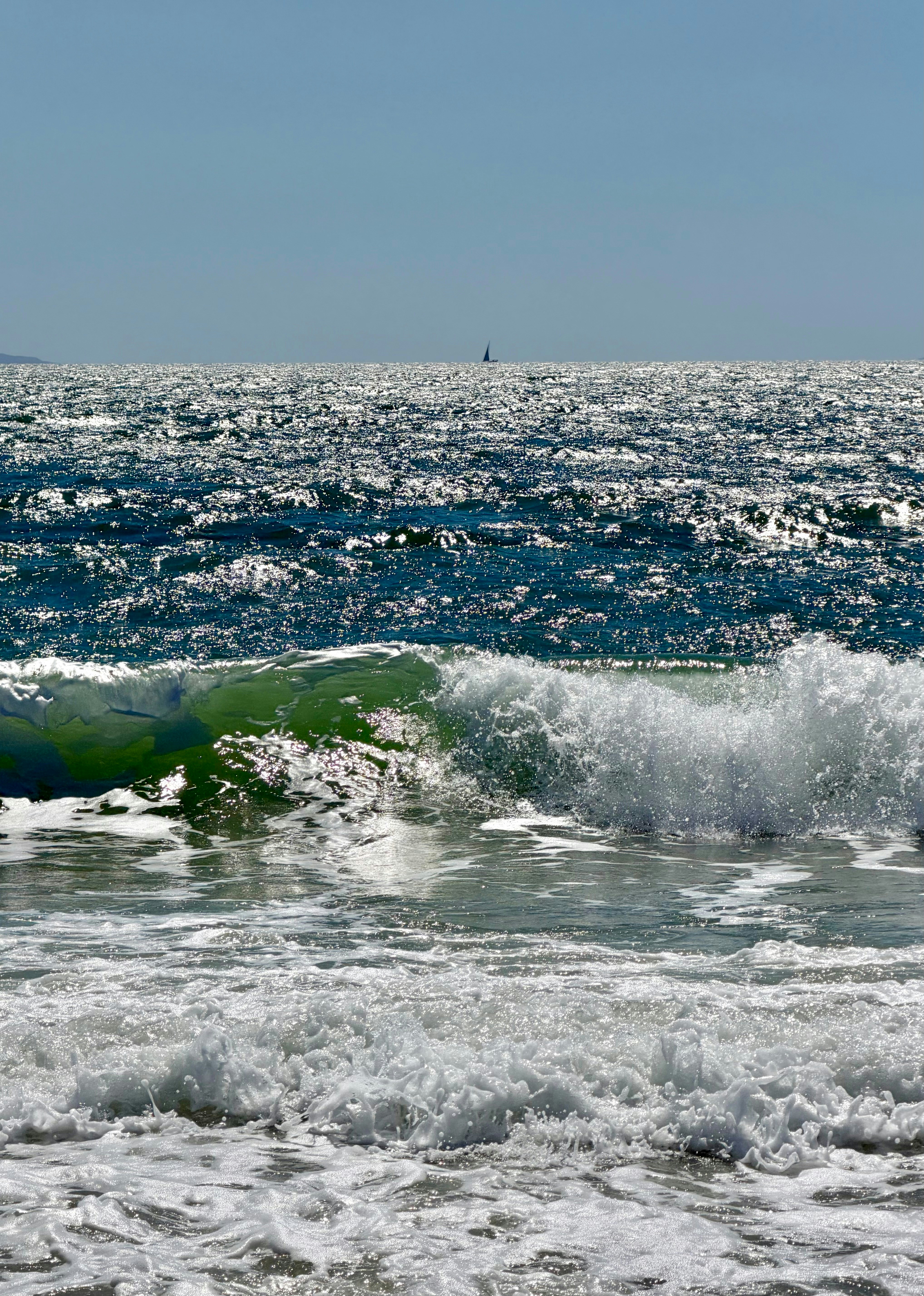 Sunlit horizon over a textured sea with foaming waves; distant sailboat silhouette near the center with sparkling water in the foreground. This is a photographic capture of the ocean, not a logo or illustration.