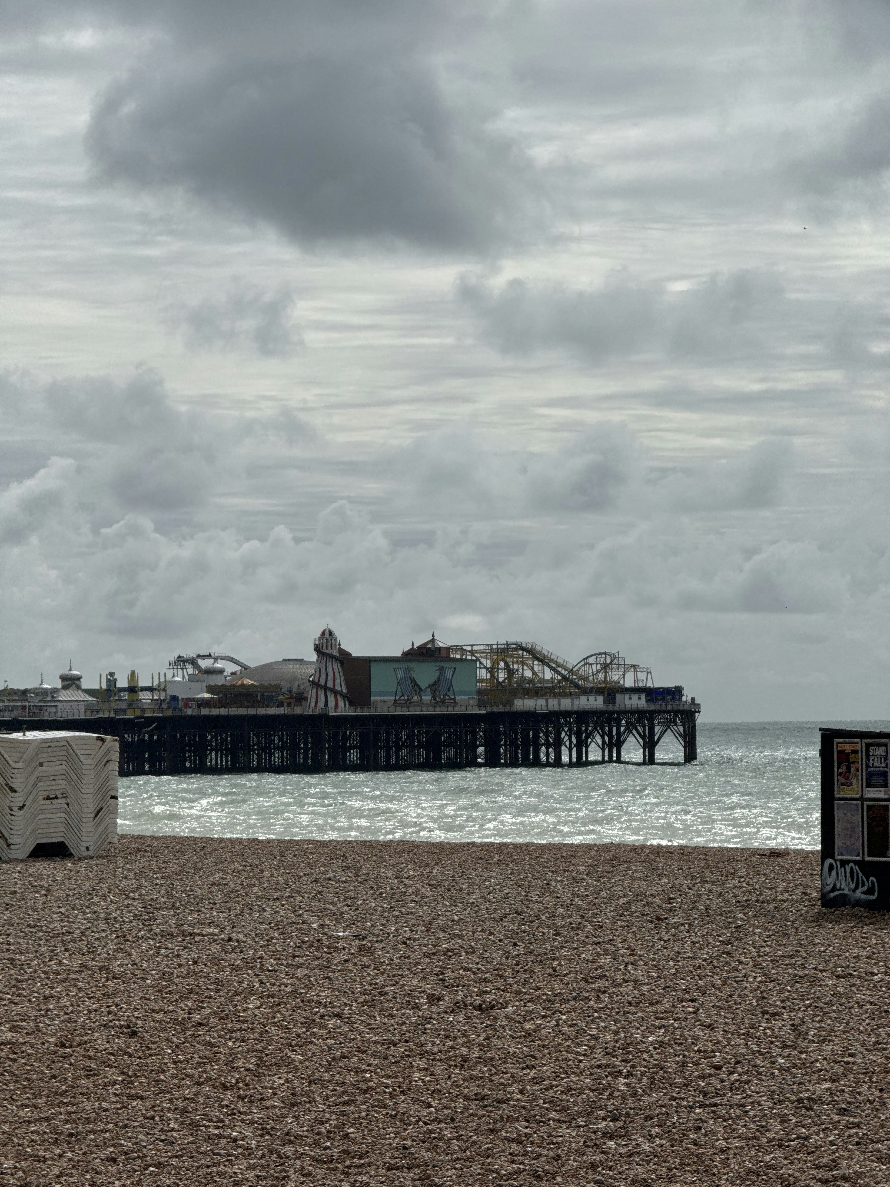Pebble beach with a distant pier and amusement structures on stilts over the sea, under a cloudy sky. A small graffiti-ed wall and stacked white beach furniture sit in the foreground.