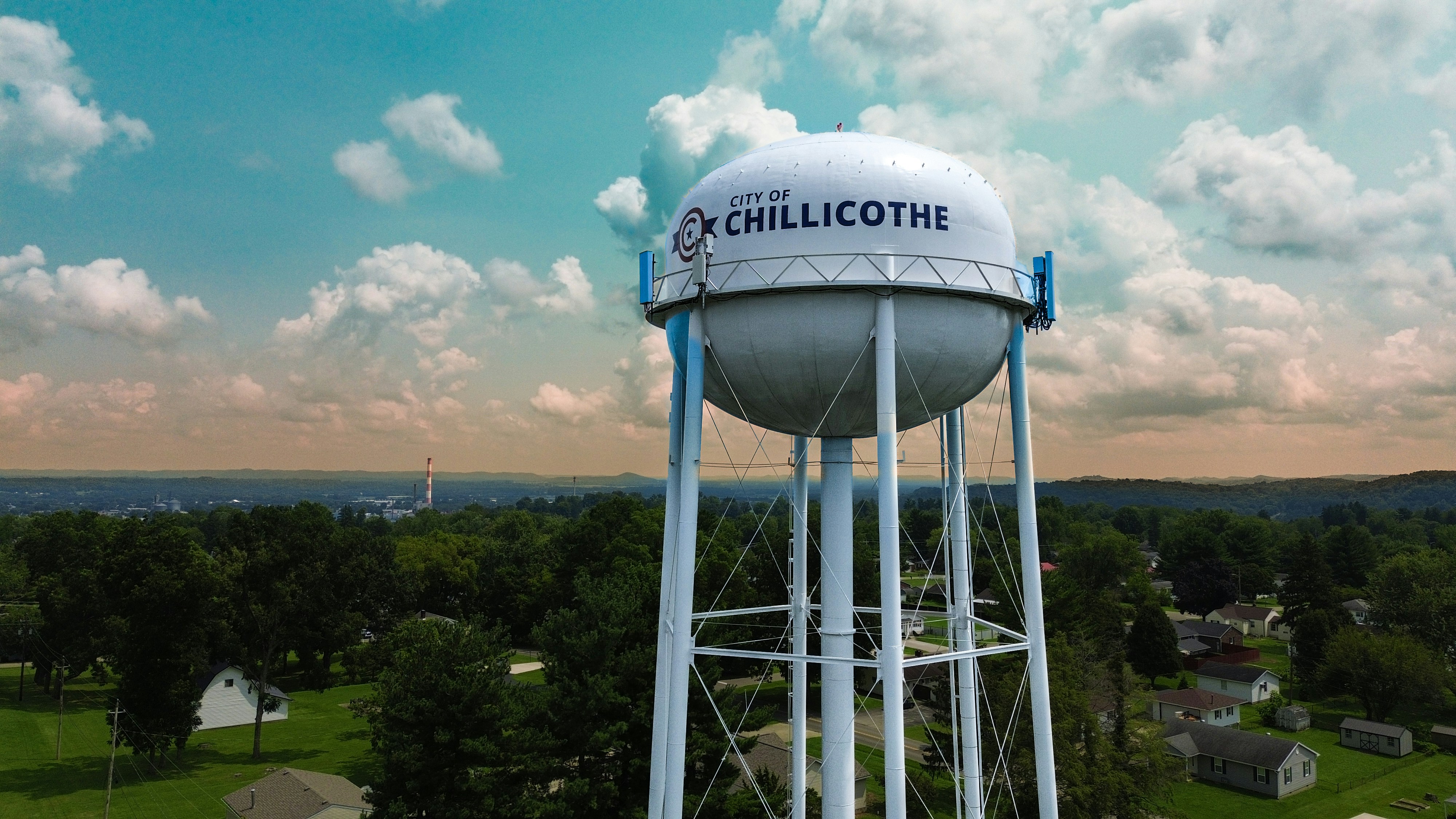 Aerial view of a white water tower labeled 'Chillicothe' rising over a small town with green fields and trees under a partly cloudy sky.