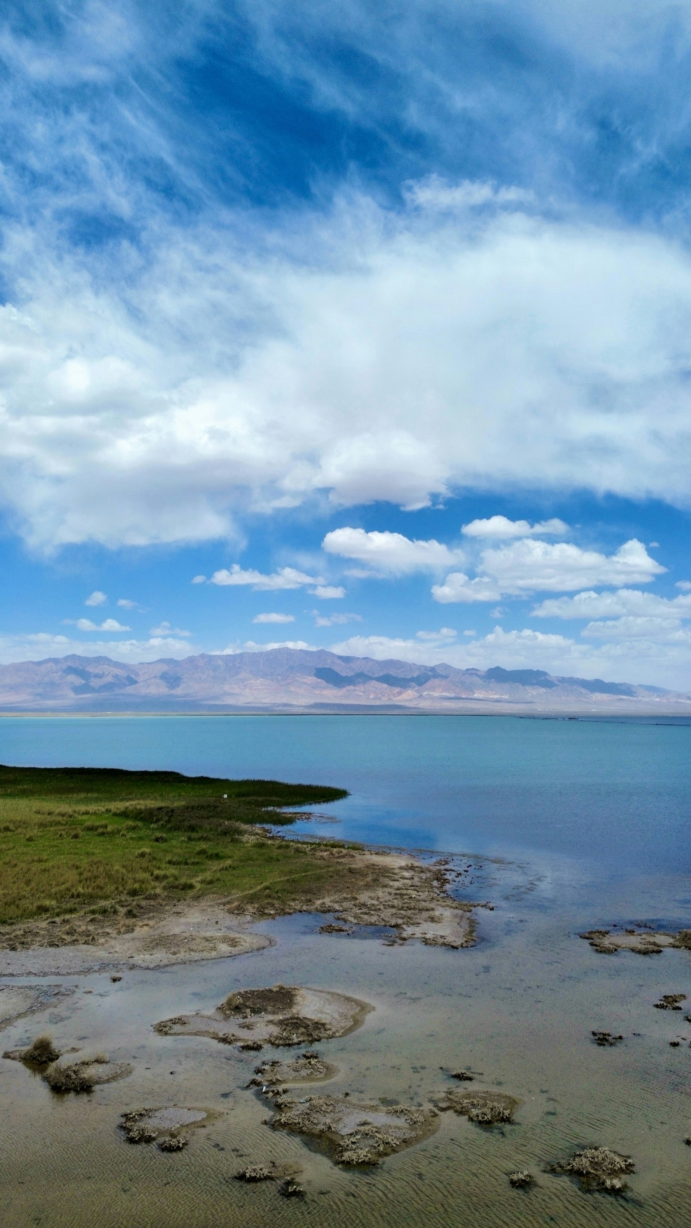 Coastal shoreline and shallow tidal pools meet calm blue water under a broad, cloud-filled sky.