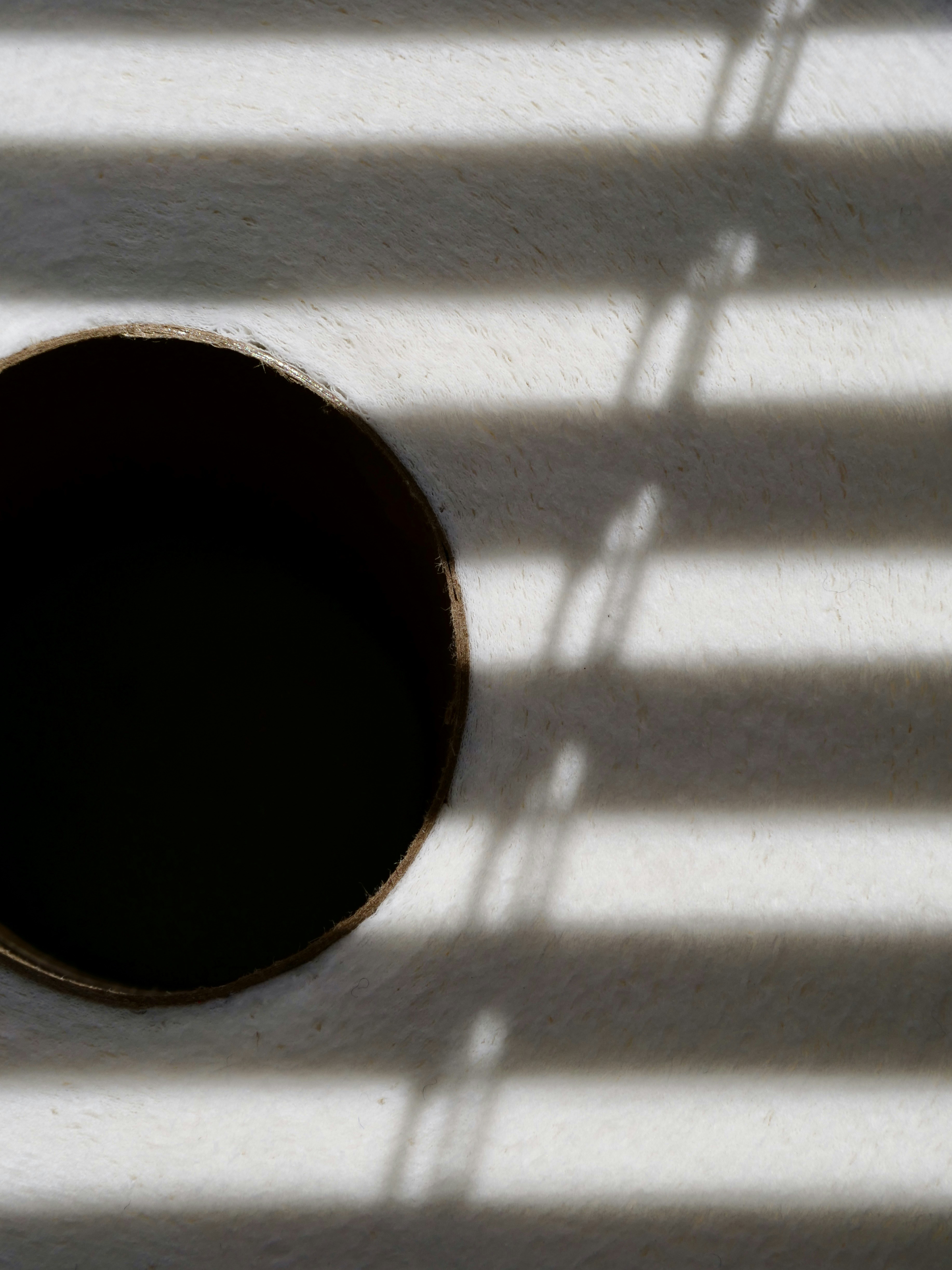 Concrete surface with a dark circular hole and horizontal shadows crossing diagonally, creating geometric rhythm. Photograph captures texture and contrast with minimal subject variation.