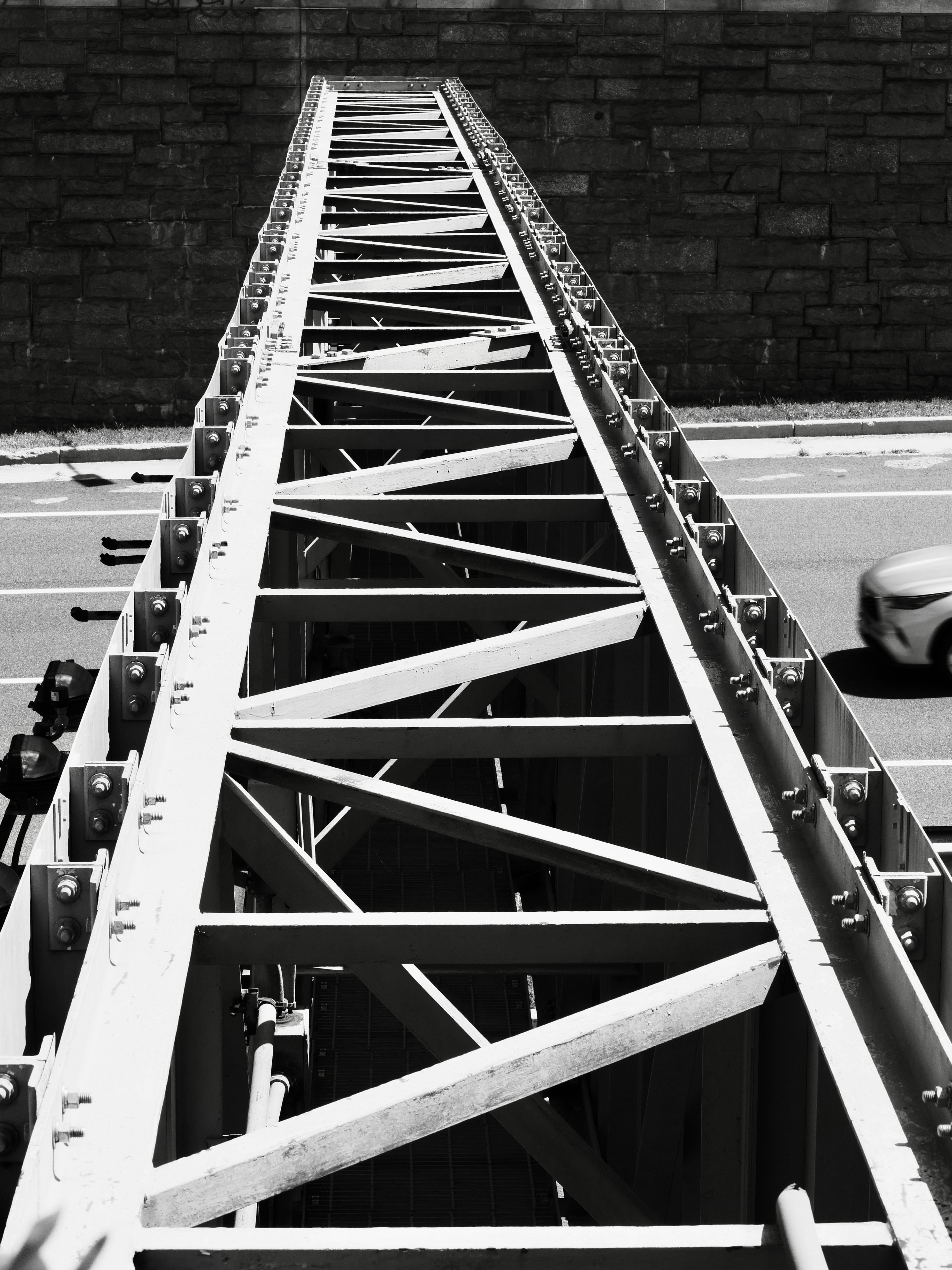 High-key black-and-white shot of a steel bridge truss rising above a street, with a moving car blurred in the background; emphasizes geometry, texture, and perspective.