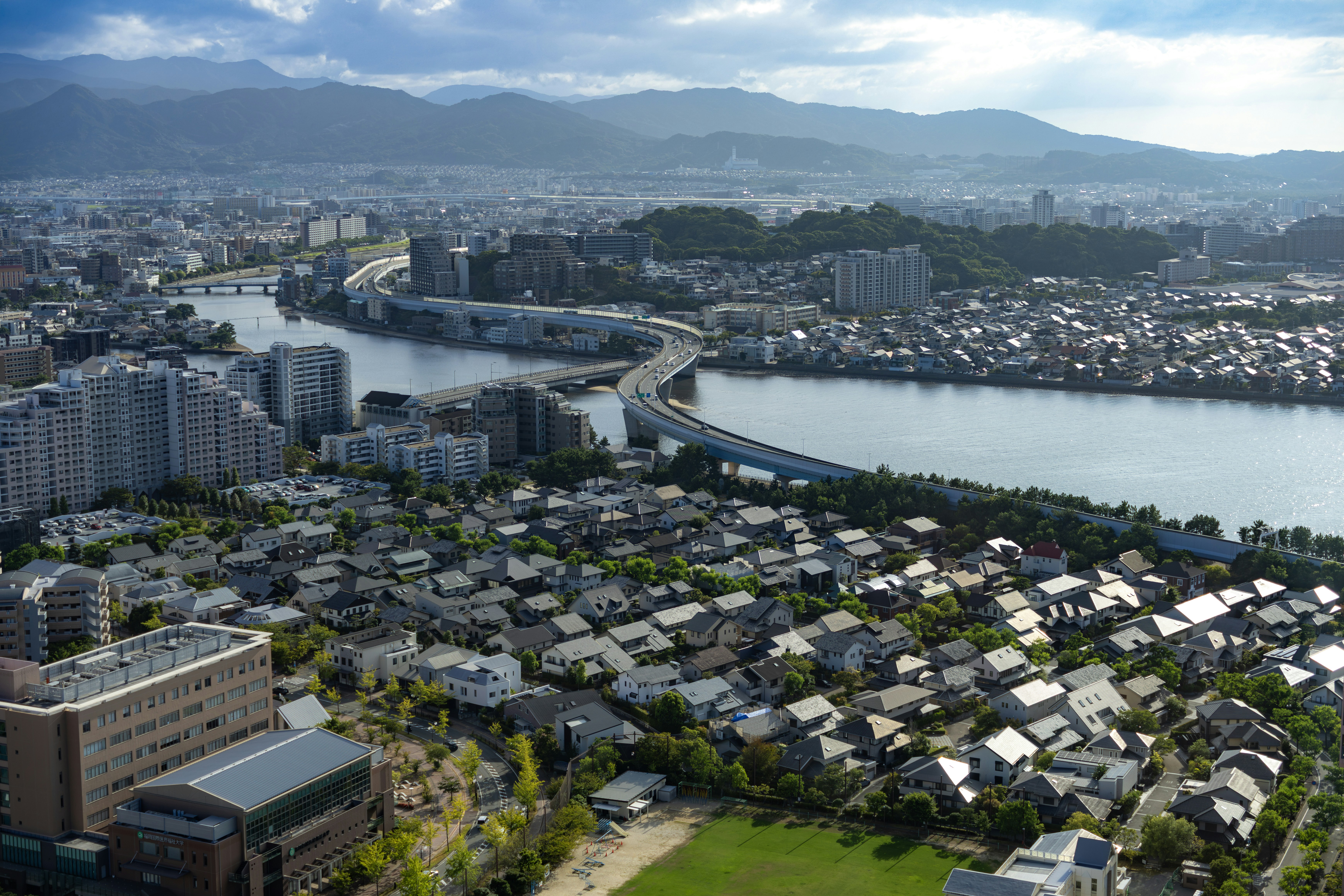 Aerial view of a city with a winding river cutting through dense residential blocks and a distant mountain backdrop. Distinct bridges and a mix of housing, with shadows and light giving depth to mid-day scenes.