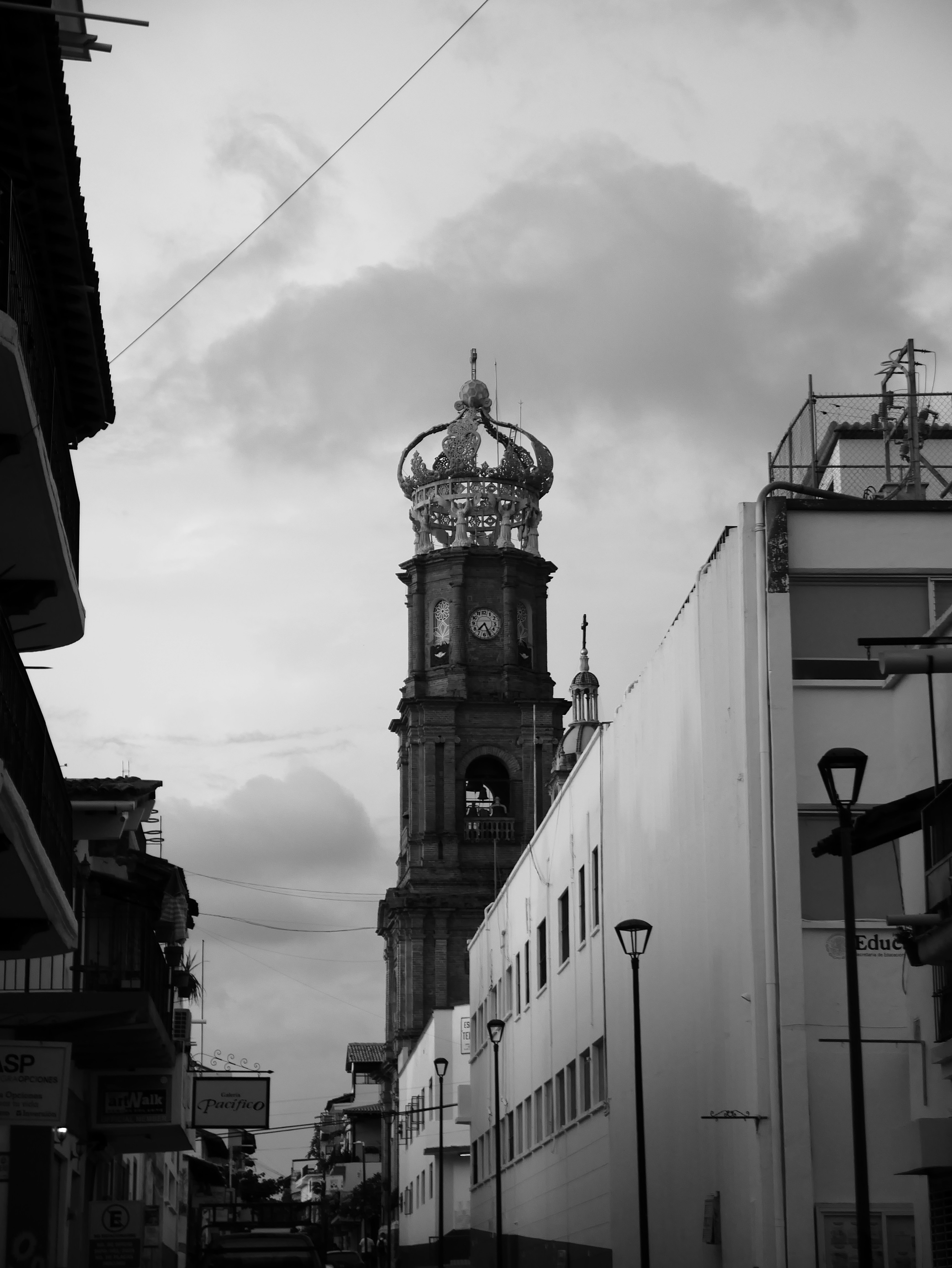 Monochrome street scene with a prominent clock tower crowned by ornate structure, rising between two rows of buildings; foreground lanes and lamp posts recede toward the horizon.