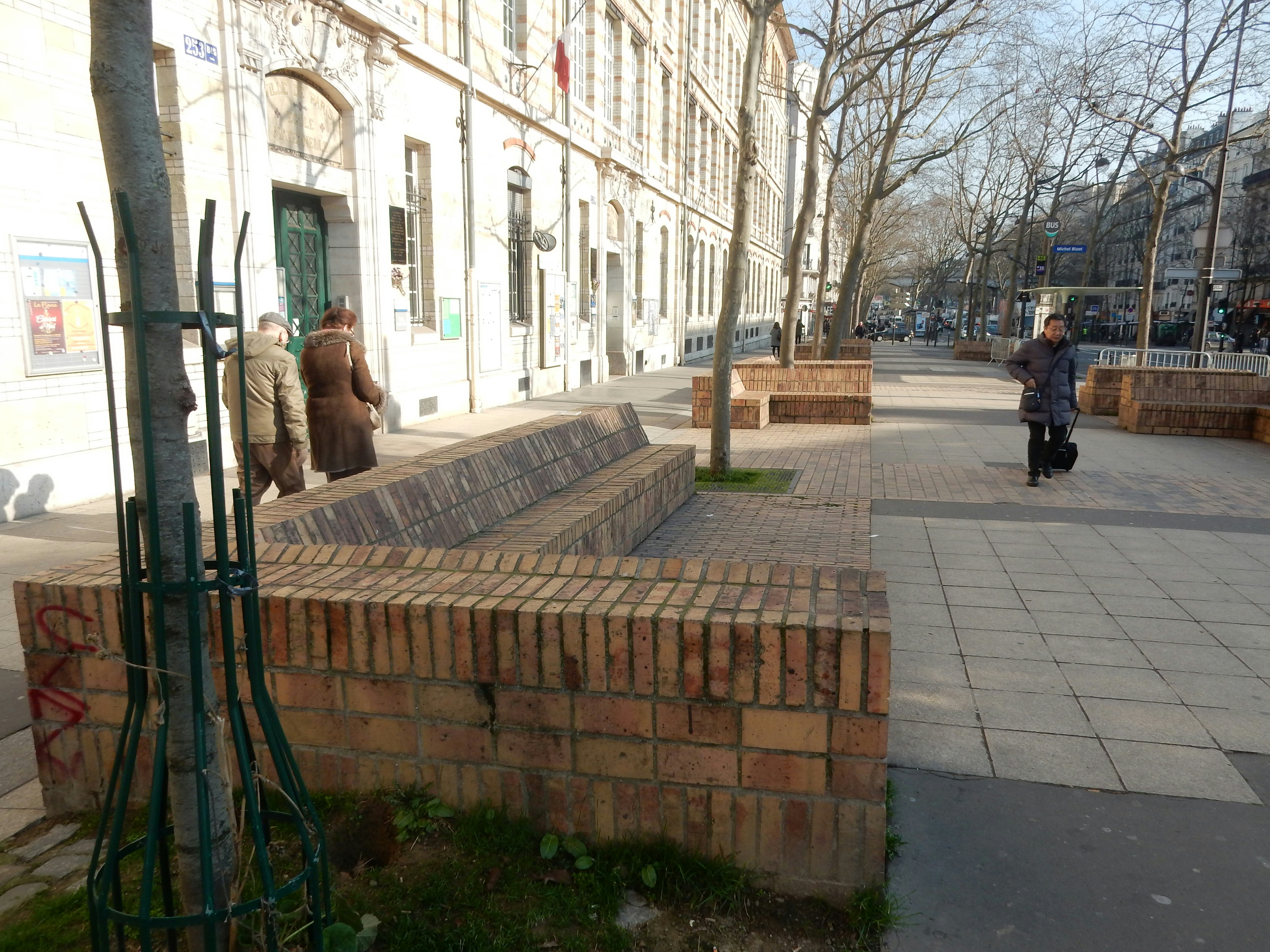 Sunlit city sidewalk with brick planters and pedestrians near a classical facade