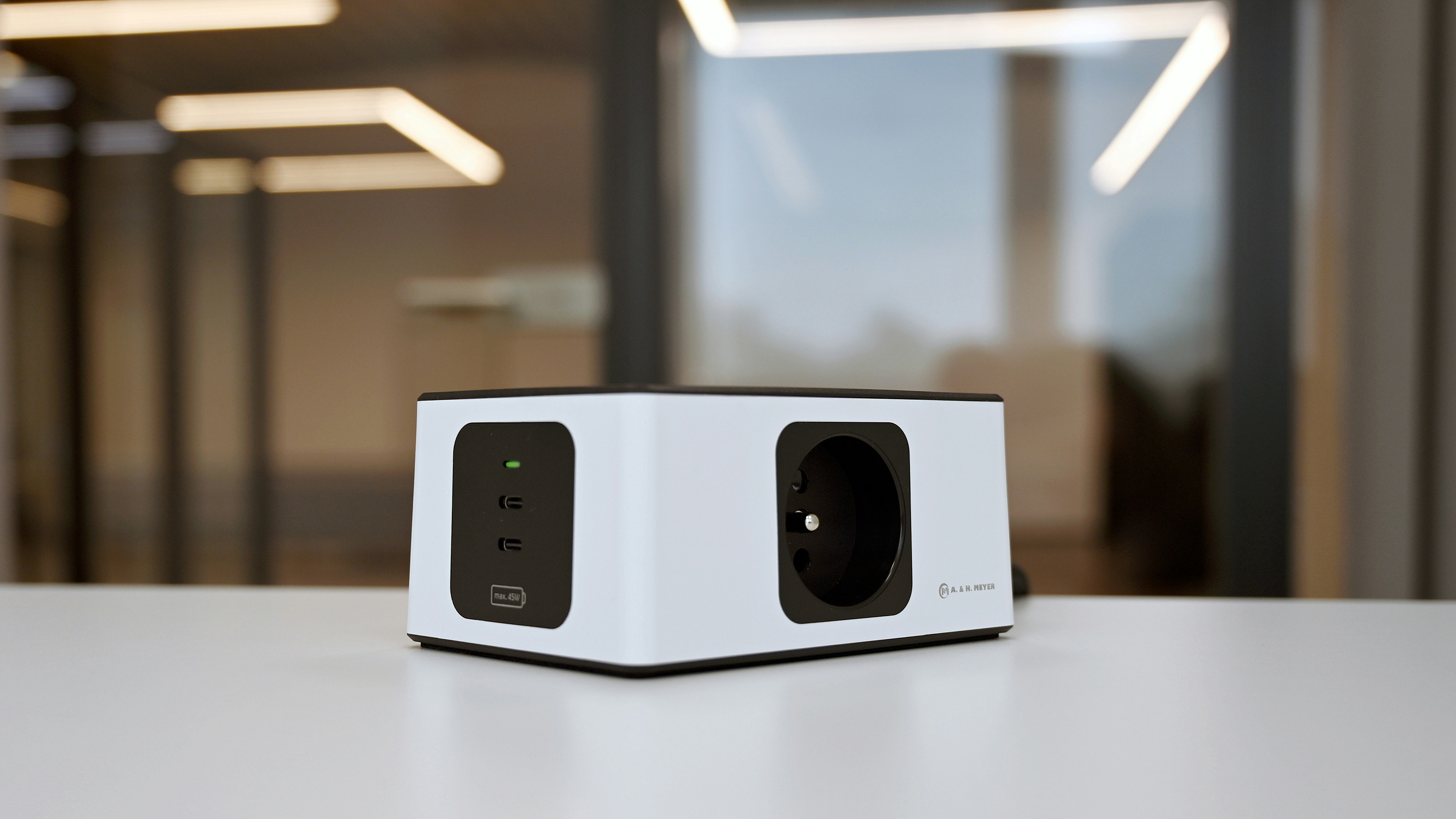 Compact white projector sits on a clean desk, its dark front panels contrasting with the bright surface; an office environment is softly blurred in the background.
