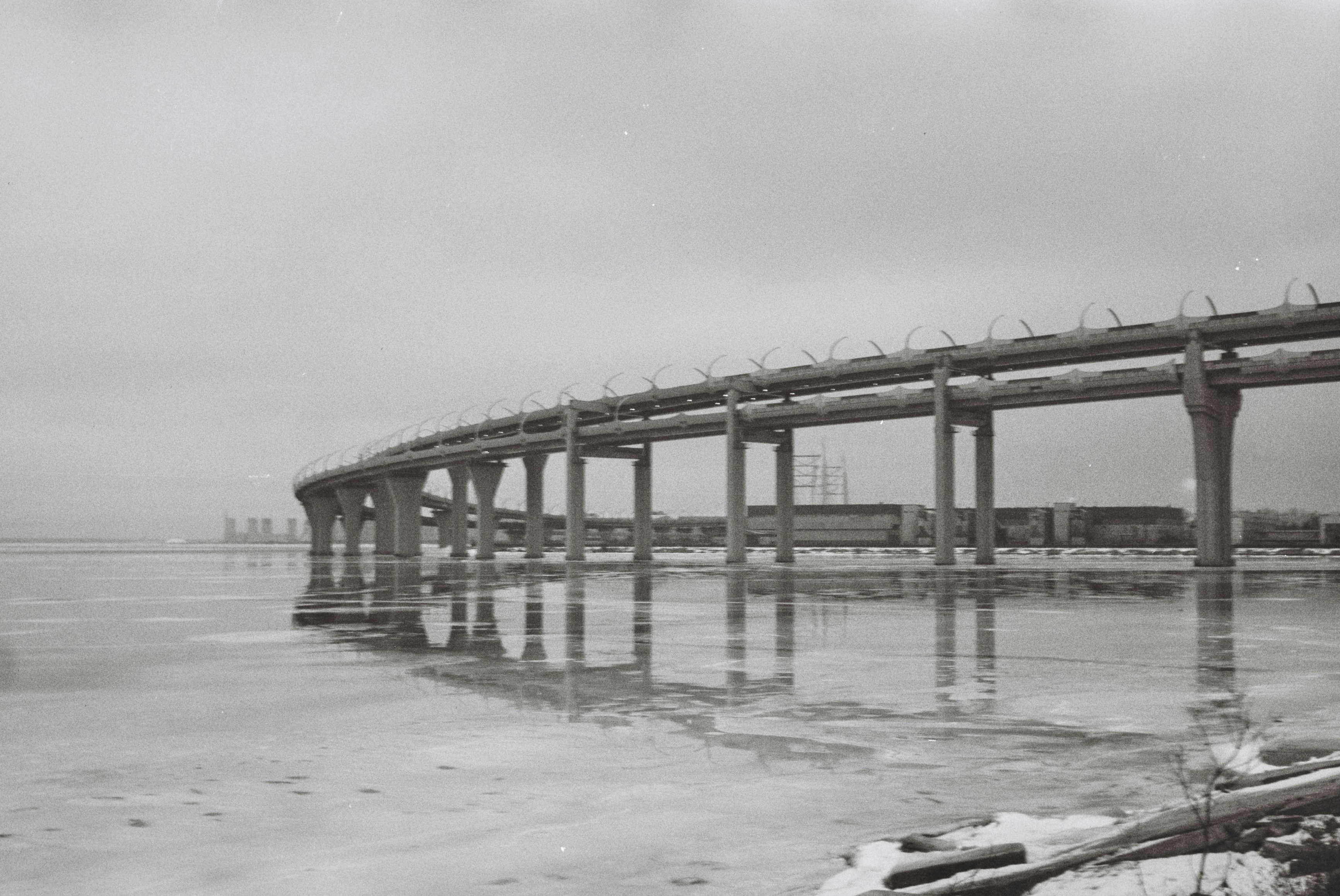 Snowy, desolate harbor with a curved bridge on stilts stretching over a frozen surface; muted tones and reflections emphasize quiet industrial stillness.