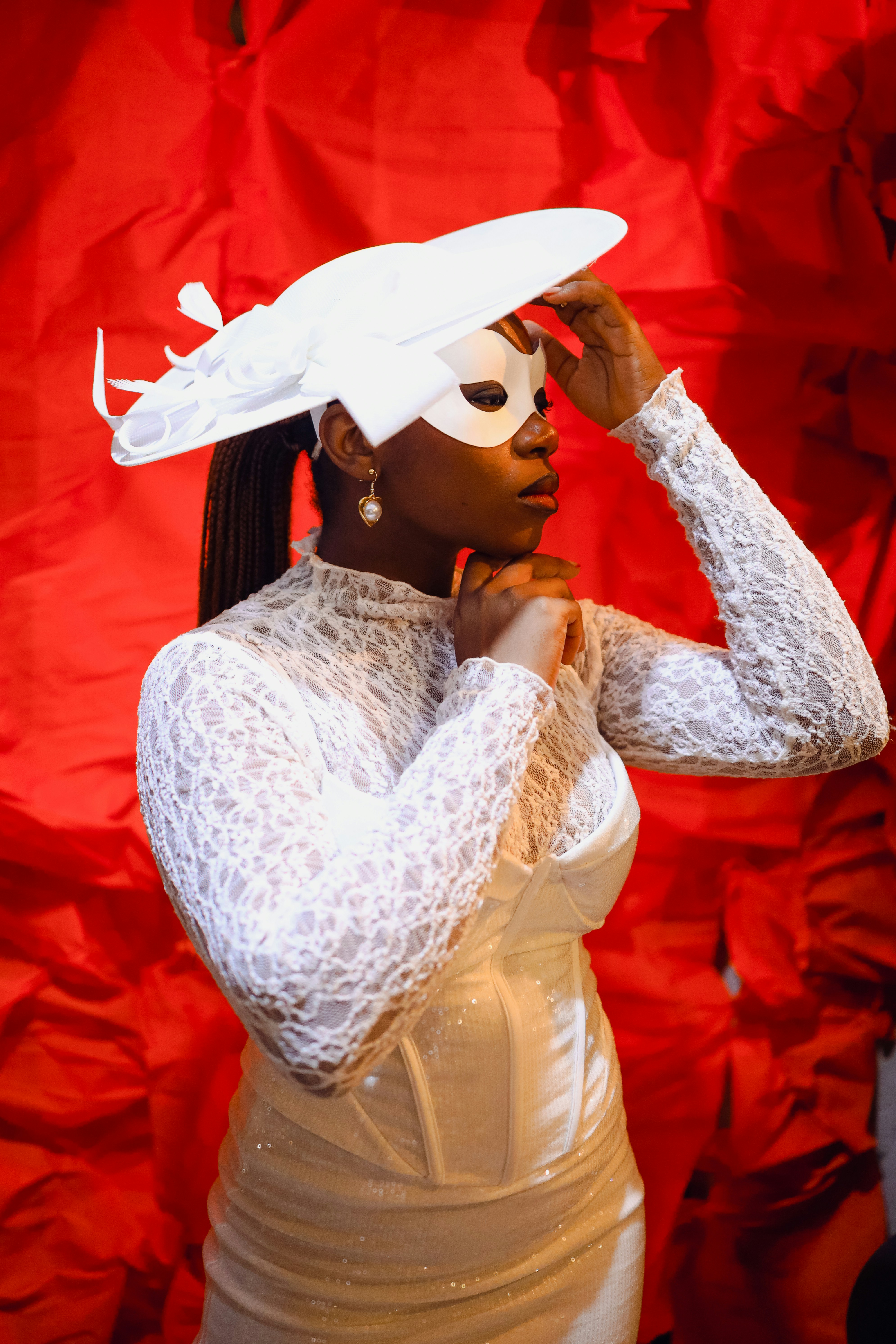 A striking fashion portrait of an elegant woman against a bold red background, wearing a sophisticated white eye mask that covers only her eyes. The contrast between the white styling and vibrant red backdrop creates a dramatic, mysterious, and luxurious editorial aesthetic.