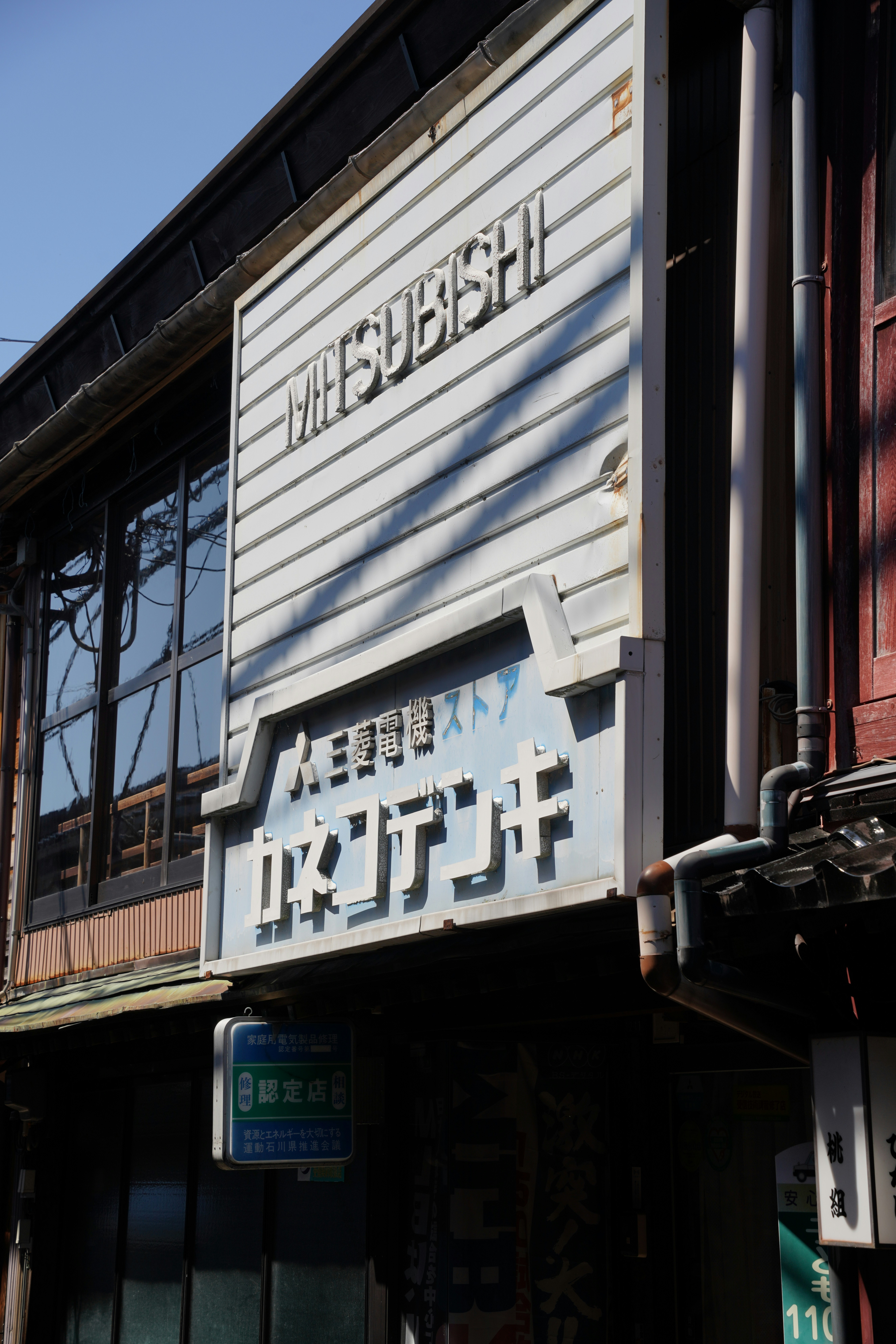 Close-up of a retro storefront sign with Japanese characters and faded white panel, under architectural framing and clear sky. A logo/advertisement sign dominates the upper portion of a building exterior.