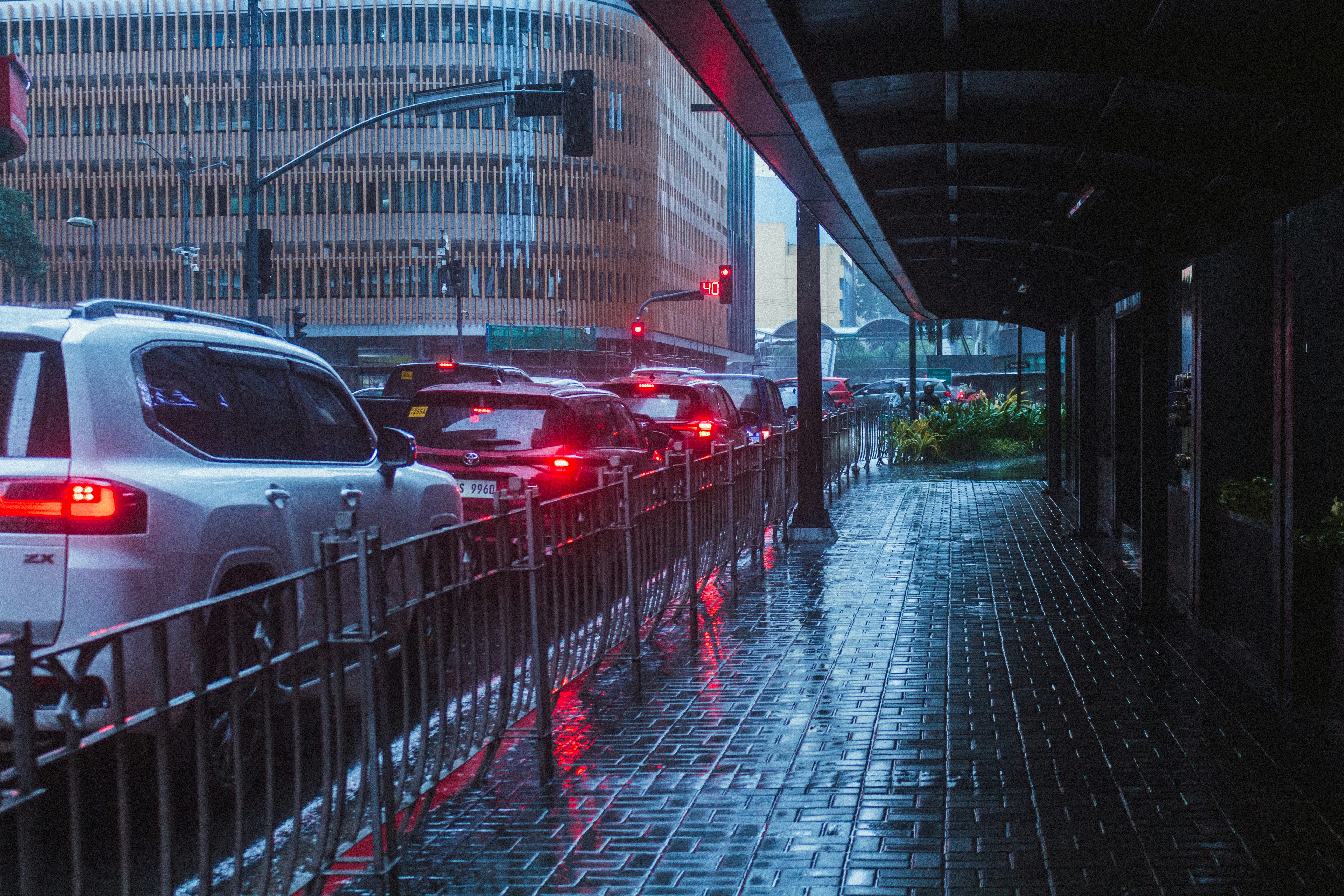 City street scene with wet reflective pavement, cars in traffic along the curb, and a covered sidewalk fading into the distance.