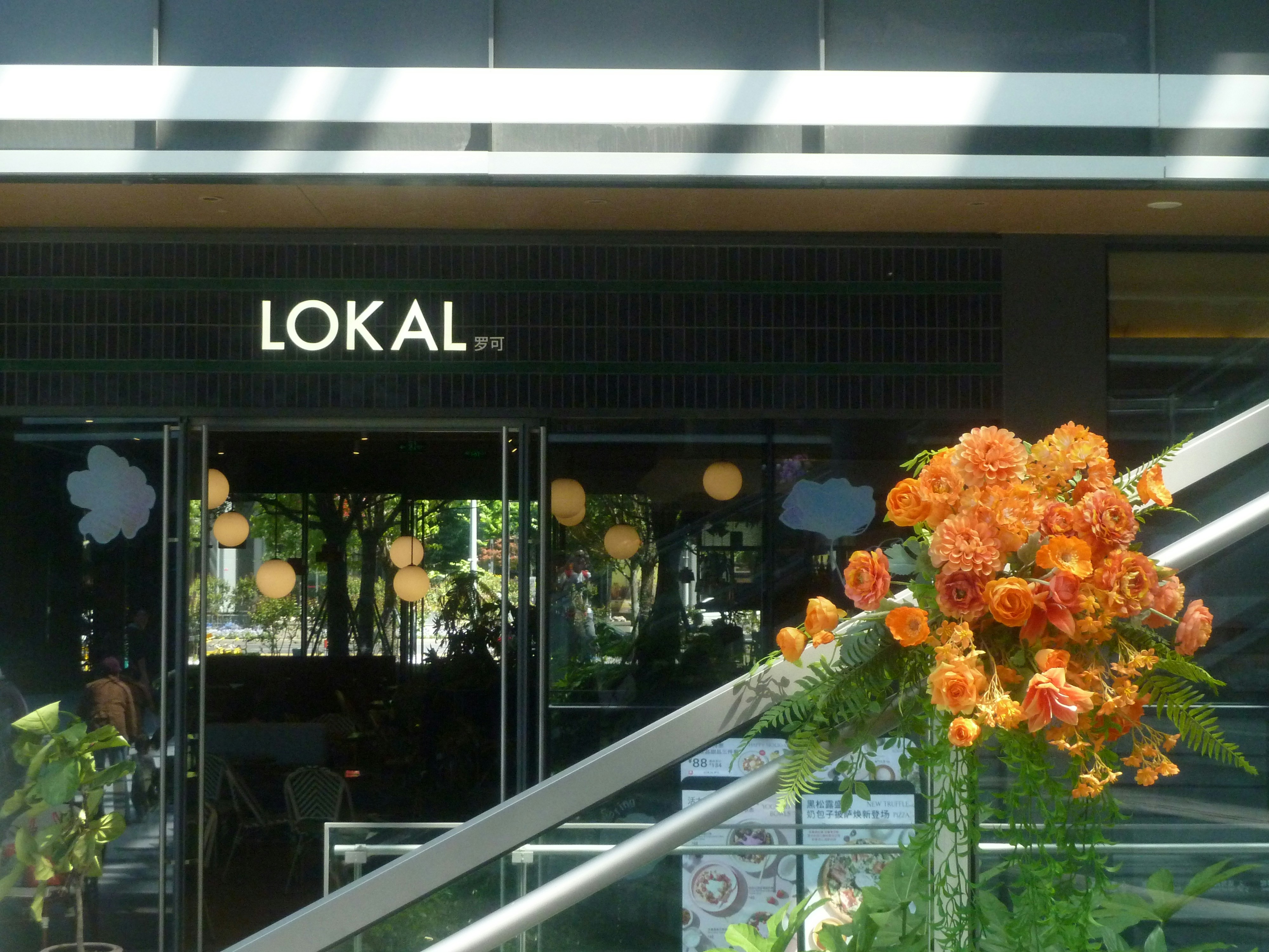 Bright storefront with 'LOKAL' sign, glass doors, and a vibrant orange floral arrangement near a diagonal railing at the entrance.