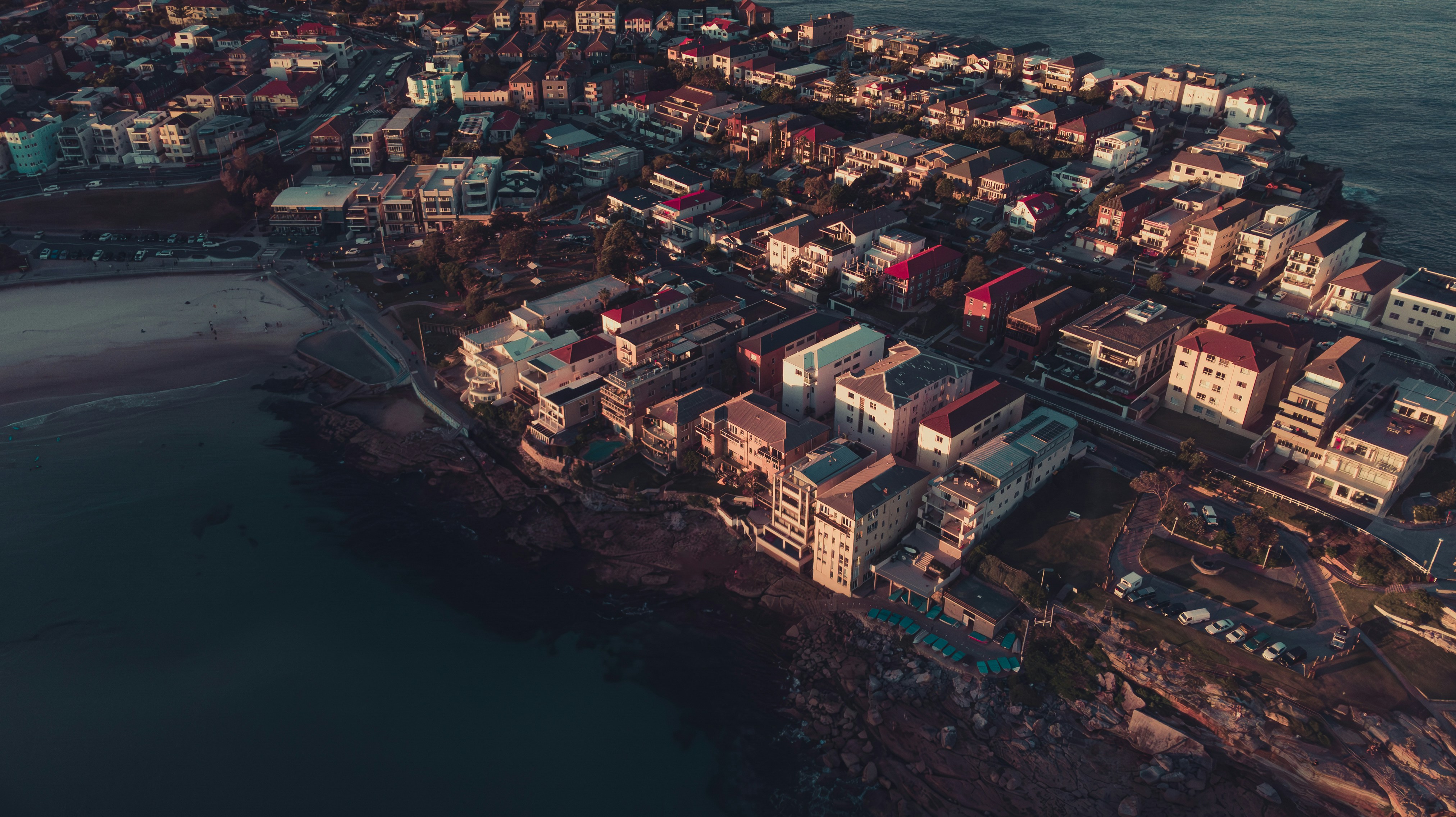 Aerial view of a densely built coastal town with multicolored buildings along the shoreline and a calm sea to the left, showcasing a compact urban grid at dusk.
