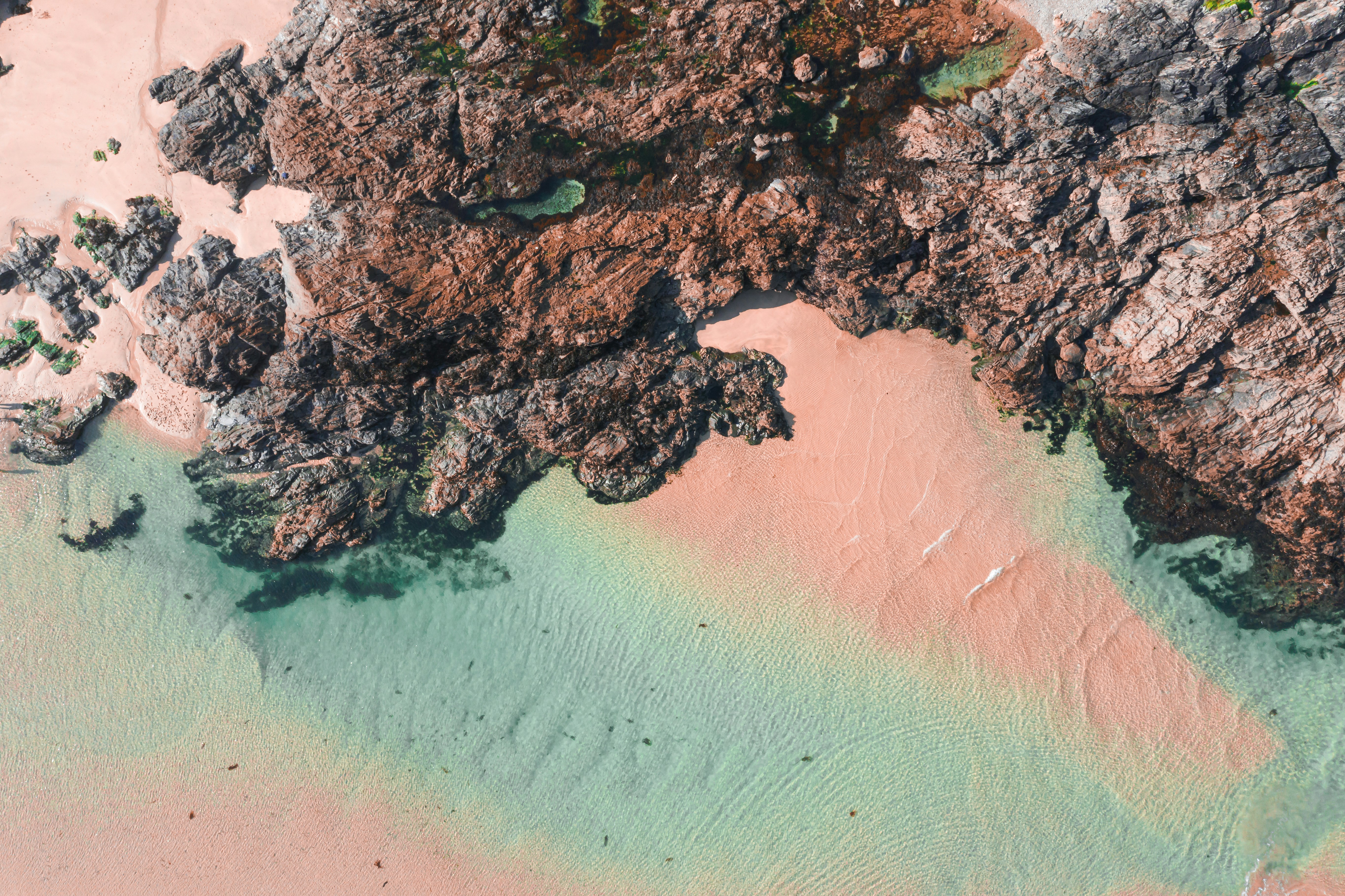 Coastal shoreline with pink sand blending into turquoise shallow water and dark rocky outcrops along the coast. Likely an aerial view highlighting contrasting textures and colors of land and sea.