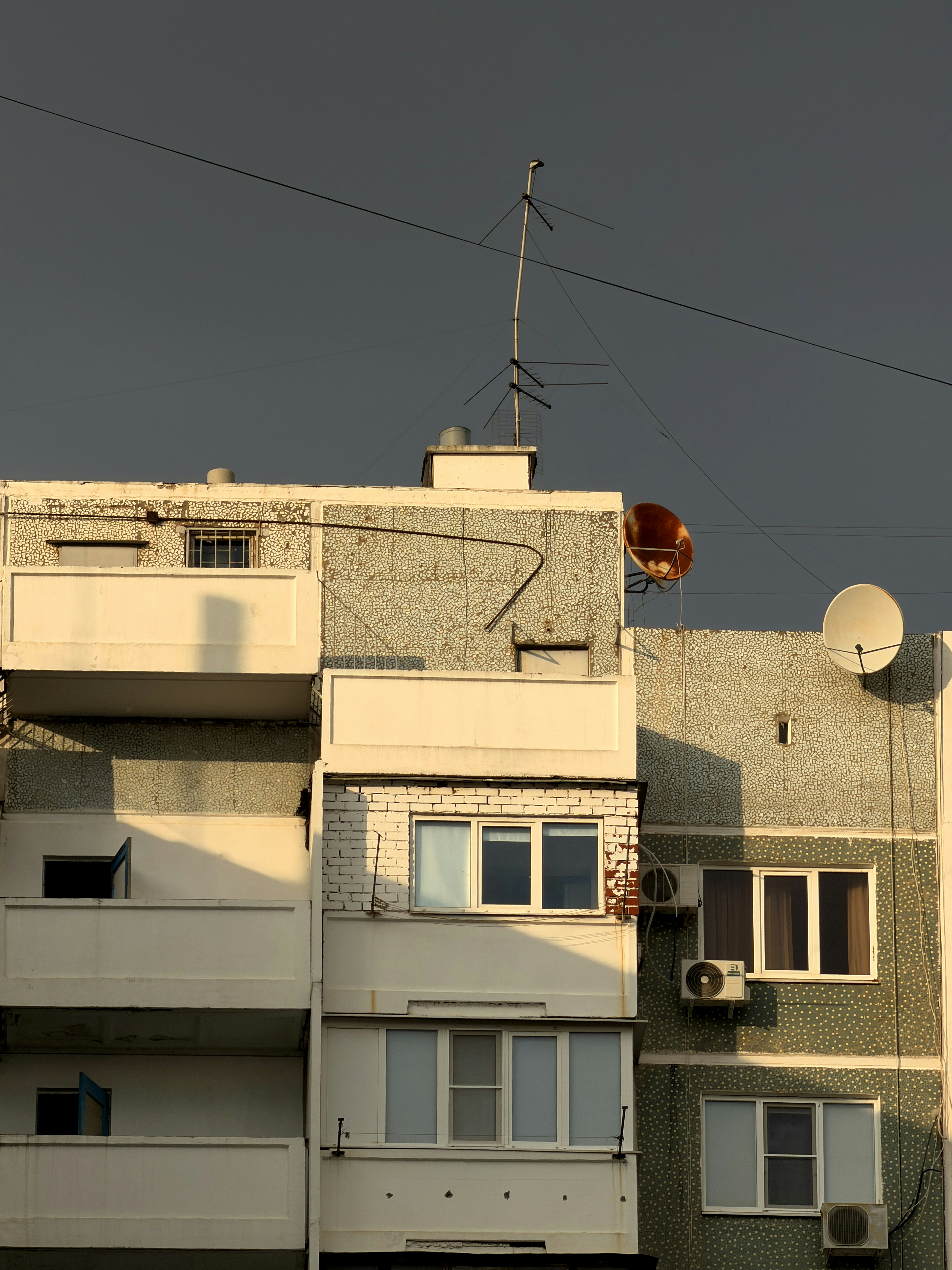 A striking architectural shot of a residential building in Krasnodar, Russia, illuminated by the warm, dramatic light of the golden hour against a dark, stormy sky. The facade exhibits a mix of textures, from mosaic tiles to white concrete balconies and brickwork, catching the intense late-afternoon sun. The roofline is punctuated by television antennas, wires, and satellite dishes, creating sharp shadows and emphasizing a post-Soviet urban aesthetic. The contrast between the bright, sunlit building and the heavy, overcast sky adds a powerful sense of atmosphere and mood.
