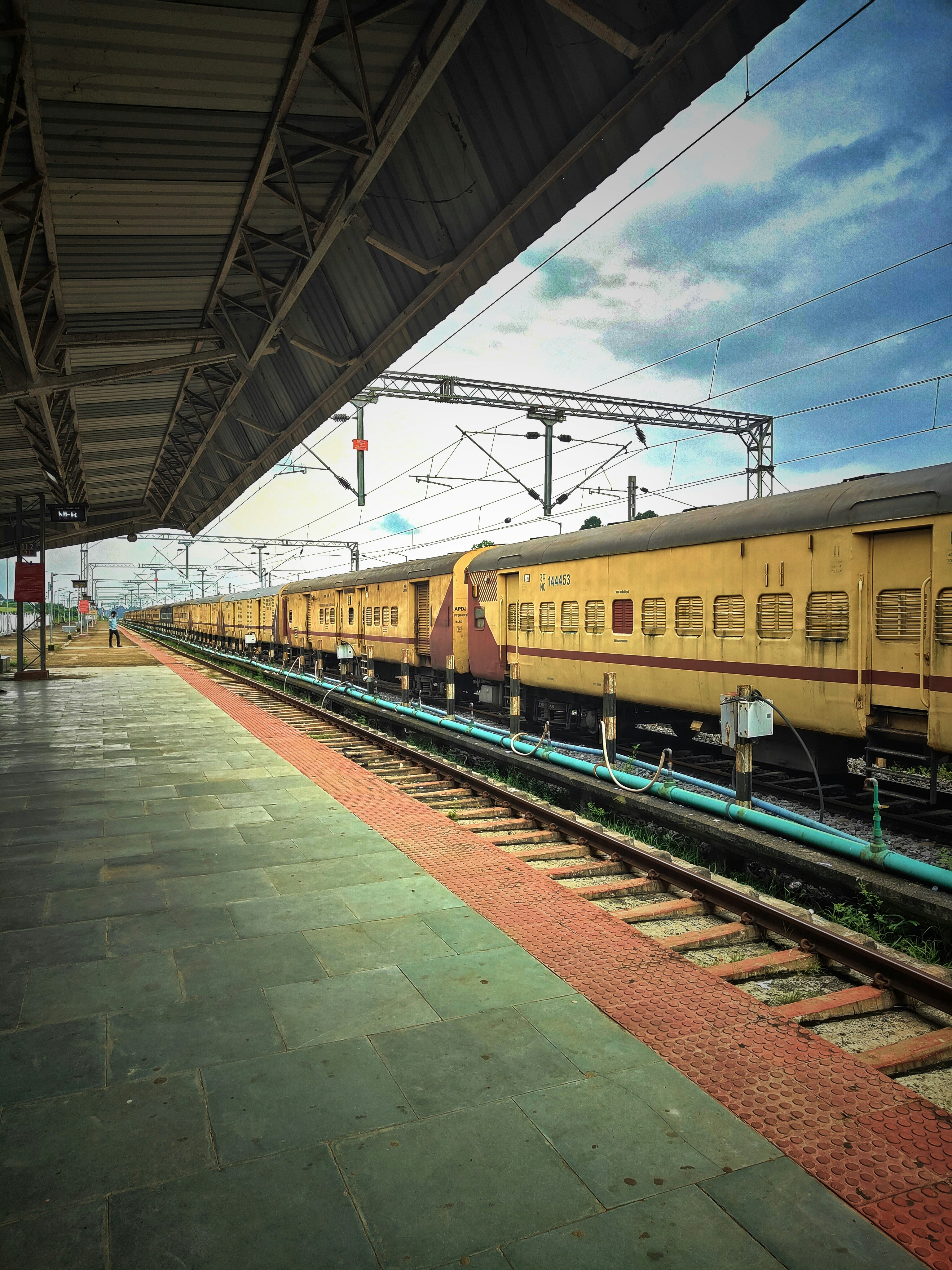 A long platform with yellow passenger cars extends into the distance beneath a steel canopy; rails and a red-tiled edge lead the eye toward distant signaling and overhead lines.