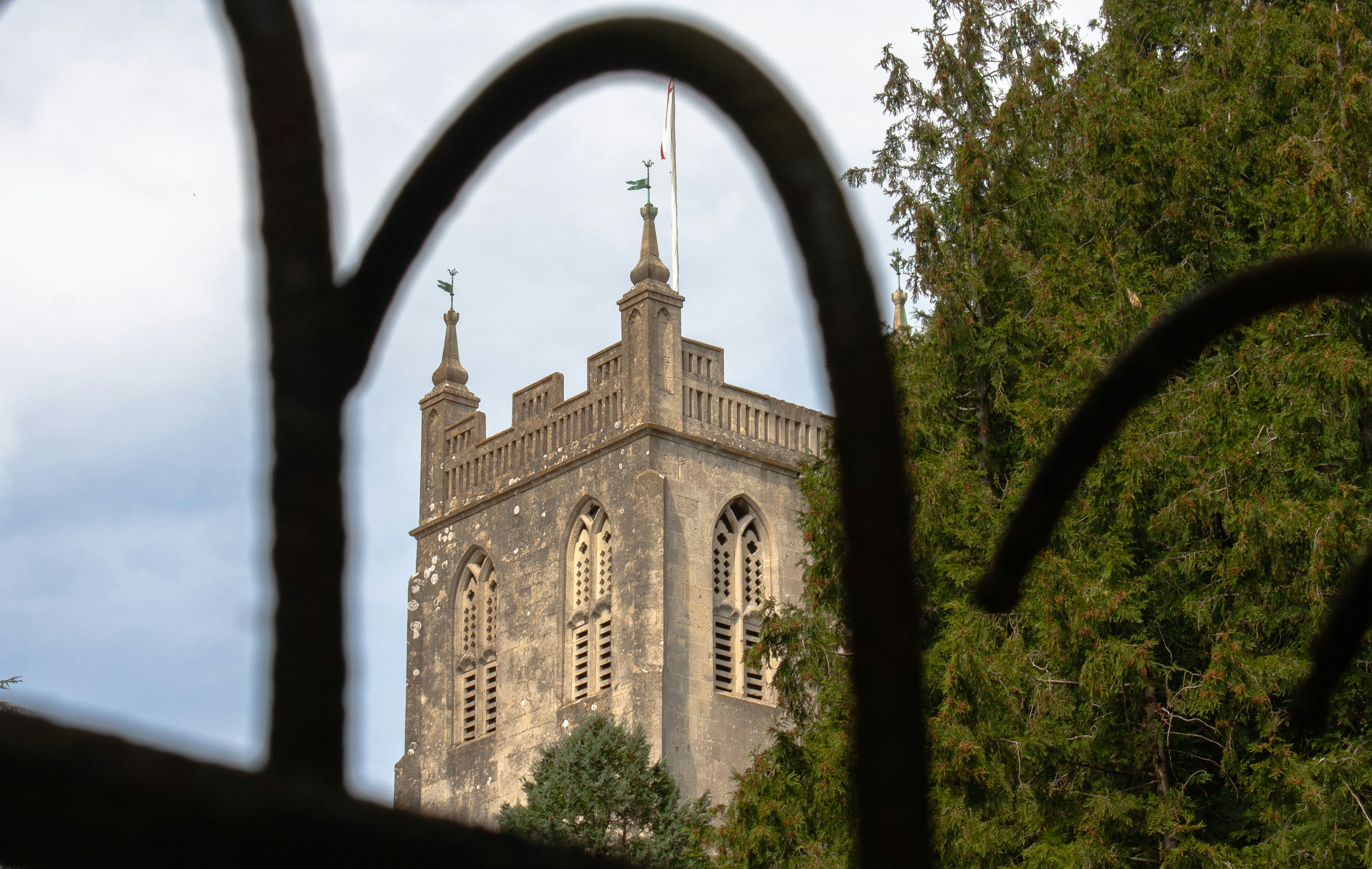 Stone cathedral tower framed by decorative wrought-iron loops with surrounding trees in soft daylight.