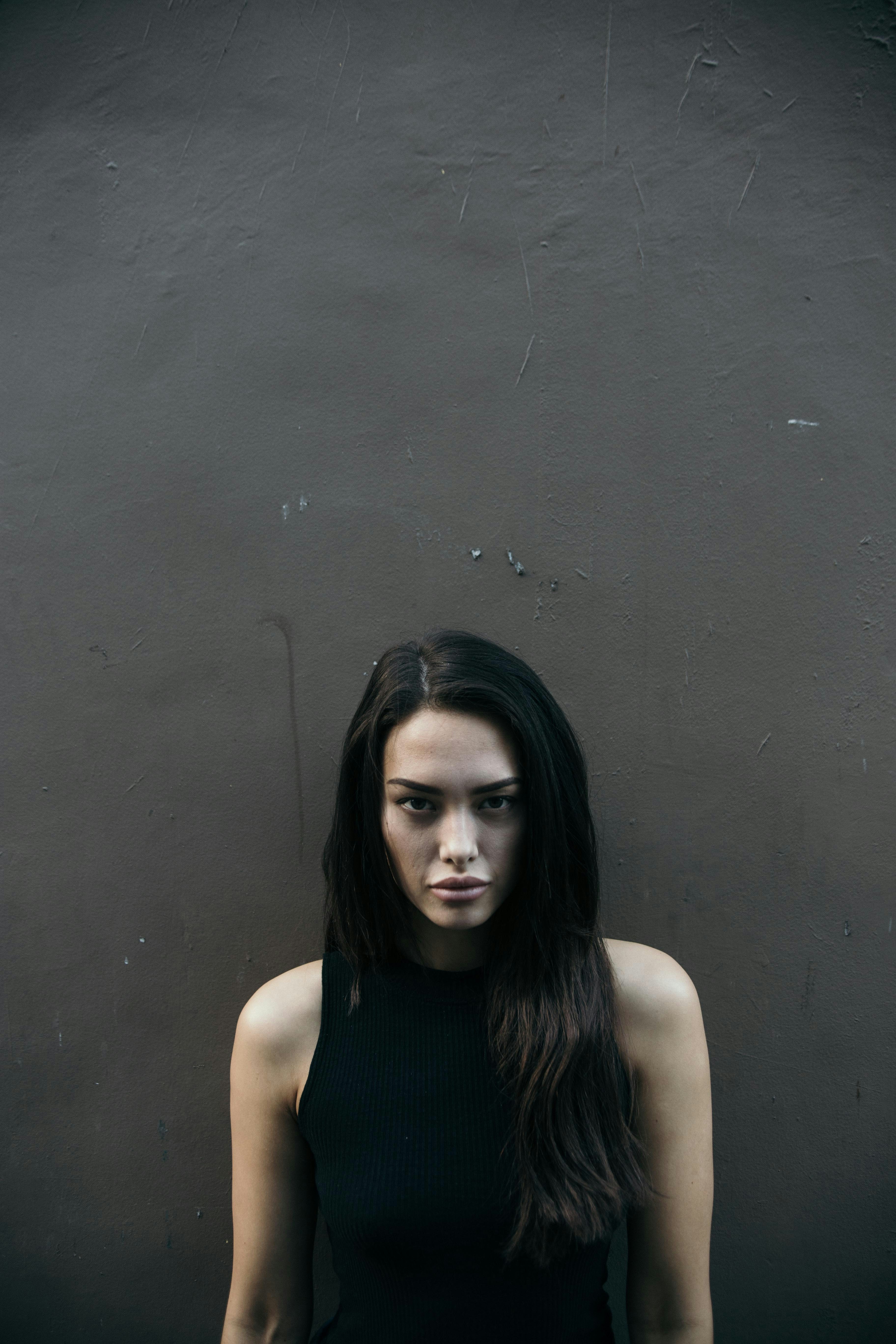 Portrait of a woman in a black sleeveless top standing against a bare, dark wall with a subdued, moody atmosphere.