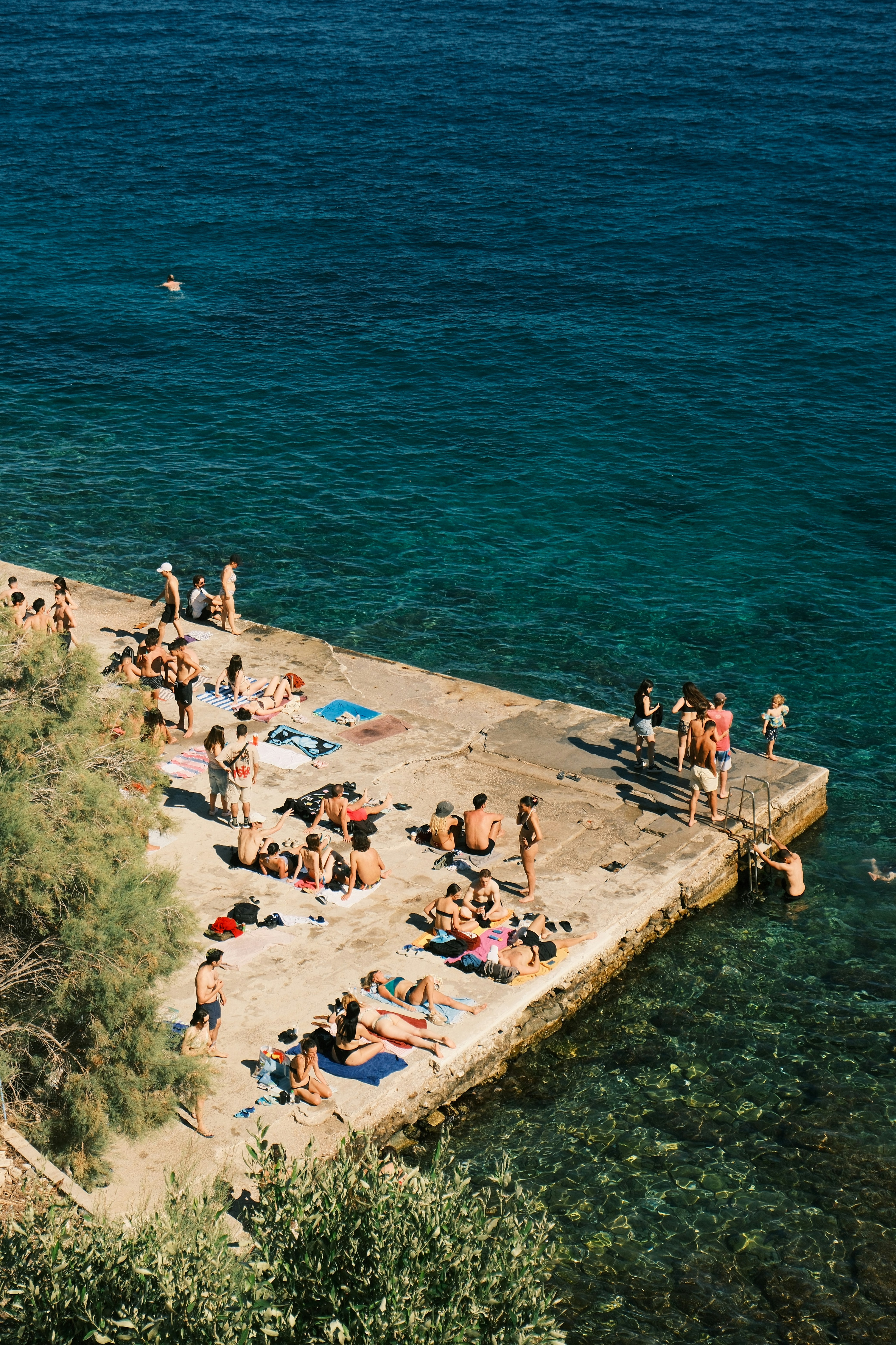 Sun-soaked beachgoers lounge on a concrete pier that reaches into turquoise waters, with the vivid sea stretching to the horizon.