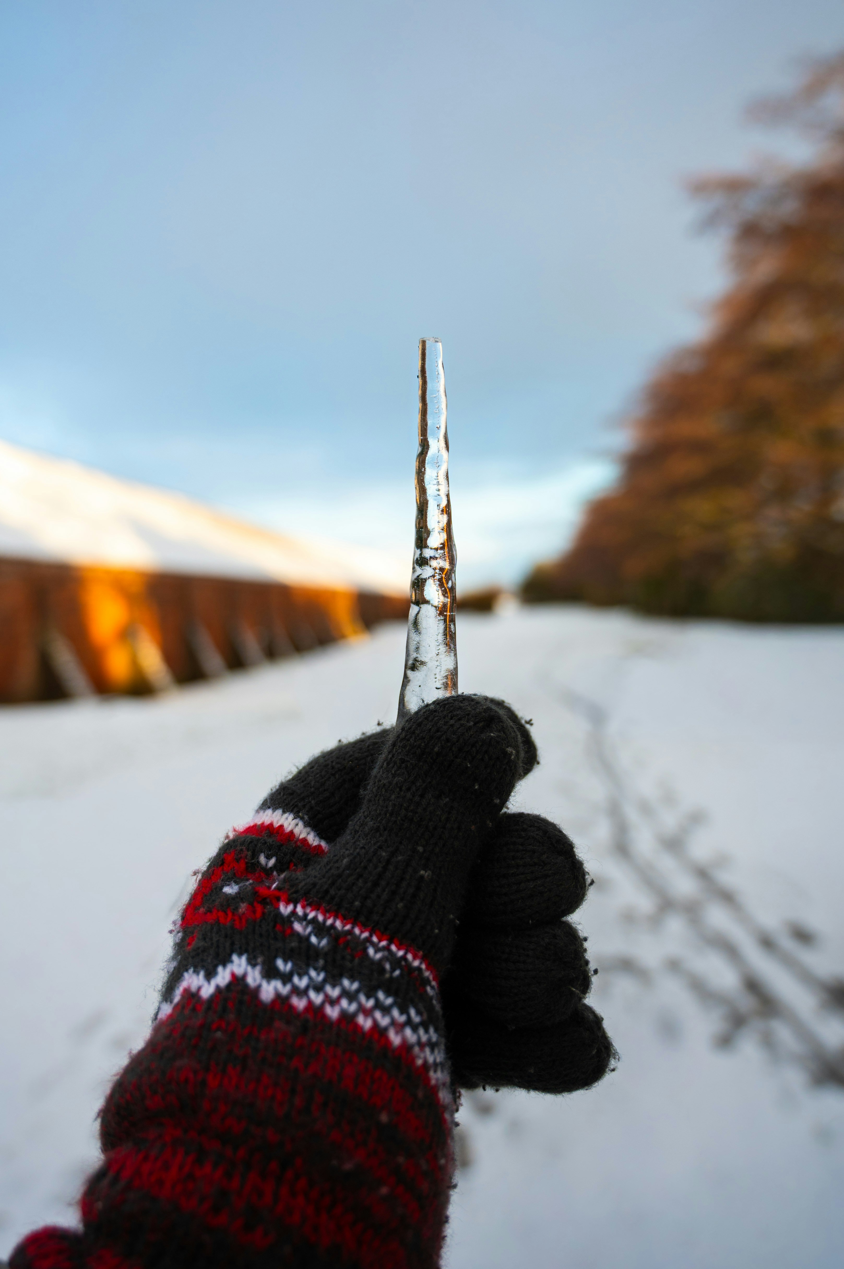 A gloved hand holds a jagged ice shard in a snowy landscape with warm-toned buildings in the background and a blue sky above. The scene emphasizes cold sensory details and a simple, focused foreground object.