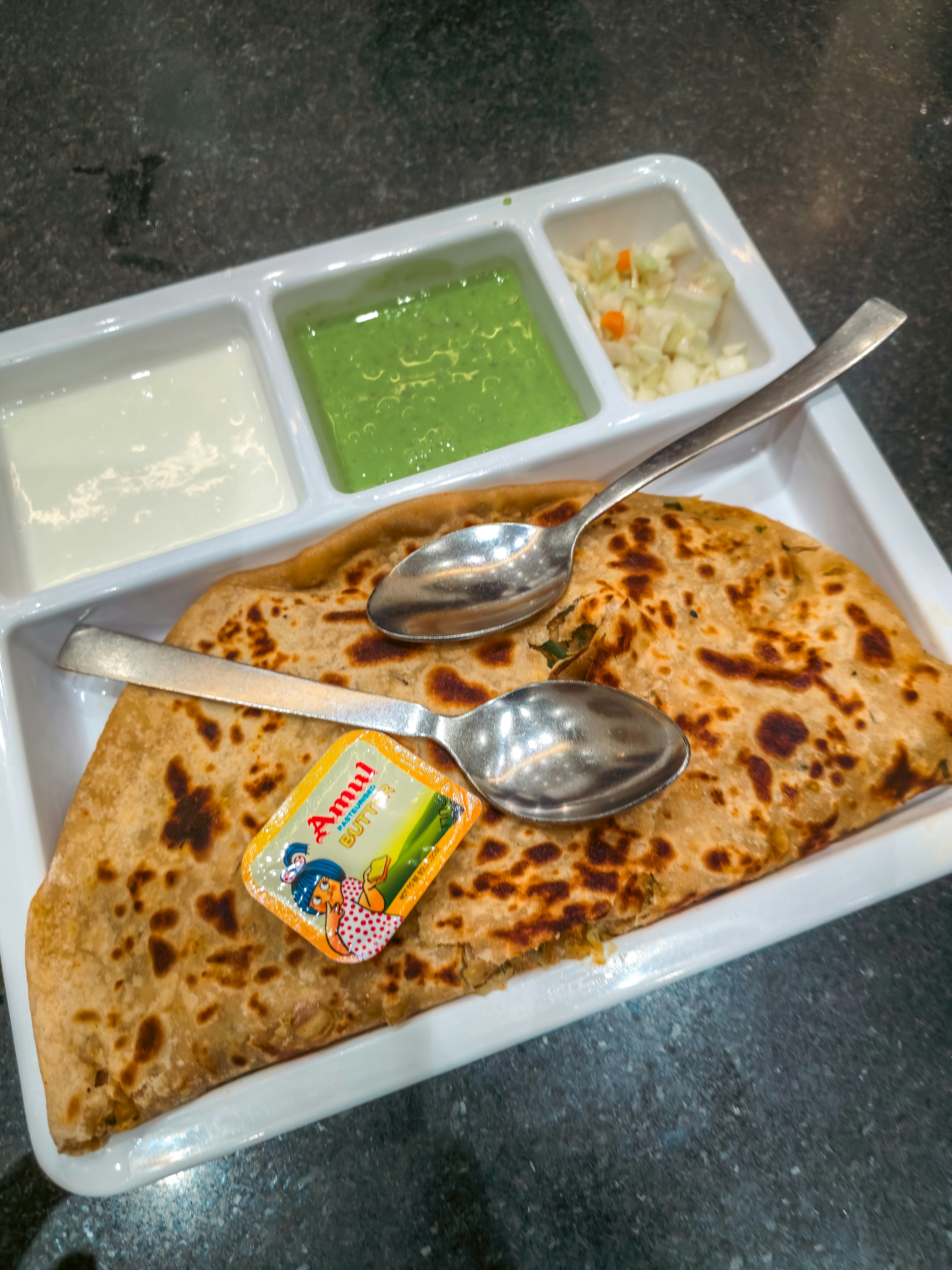 Paratha served on a segmented tray with three dipping sauces and a small butter packet, a spoon and knife resting atop the flatbread; a compact breakfast plate capturing a casual dining moment.
