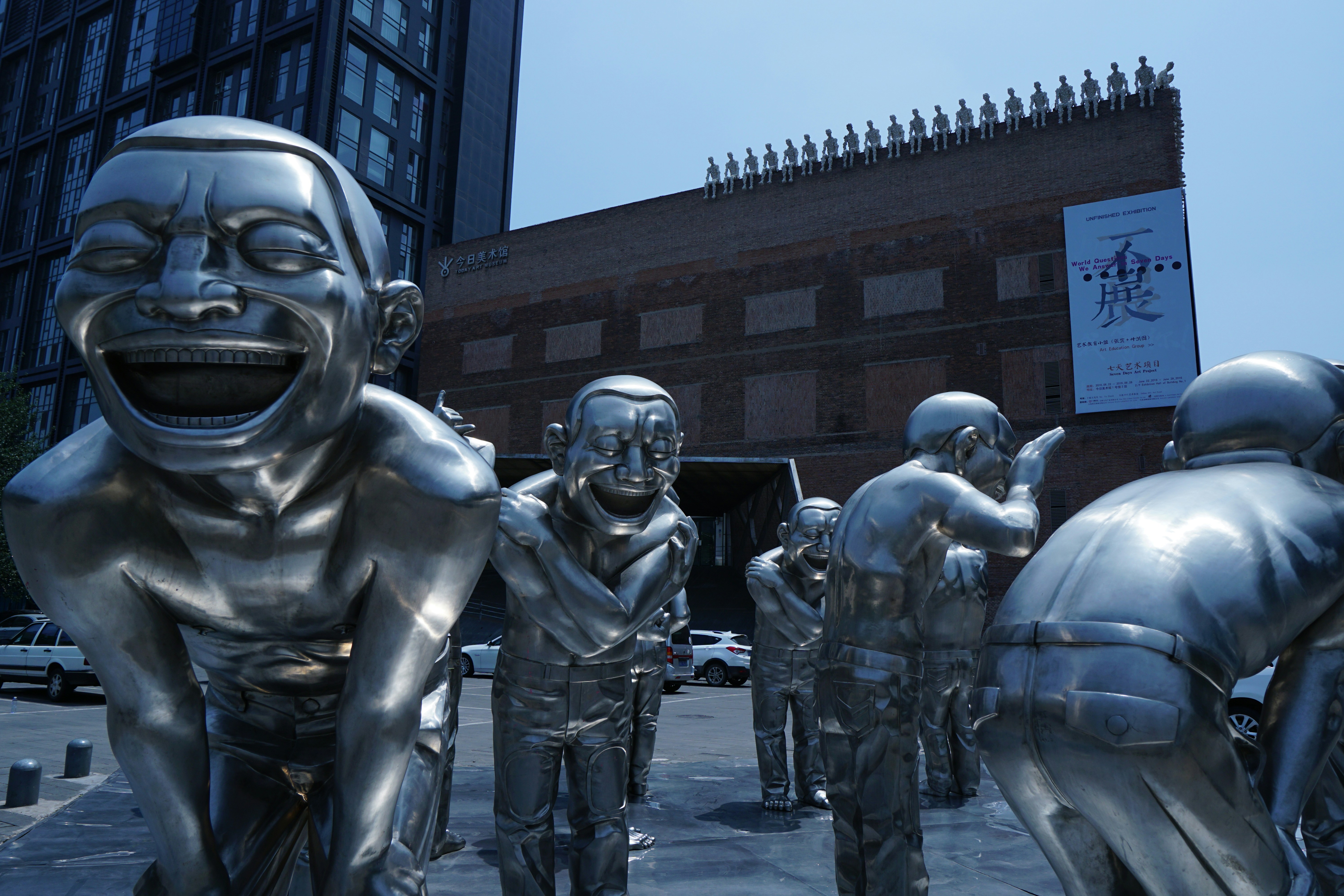 Shiny metallic statues form a row in an urban plaza, with a brick building and banners in the background, emphasizing the surreal, public-art installation.