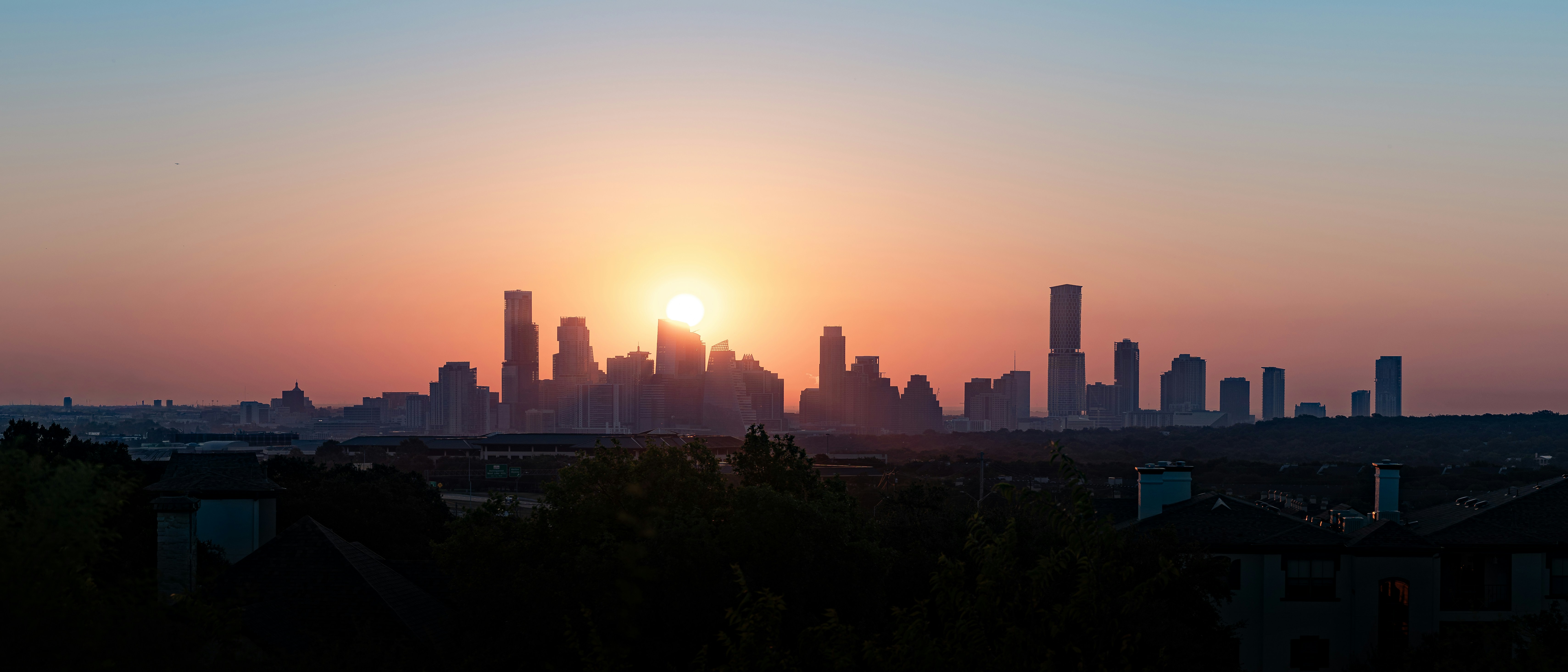 Sunset silhouettes the city skyline with a low sun just above the horizon, casting warm hues across the buildings and foreground trees.
