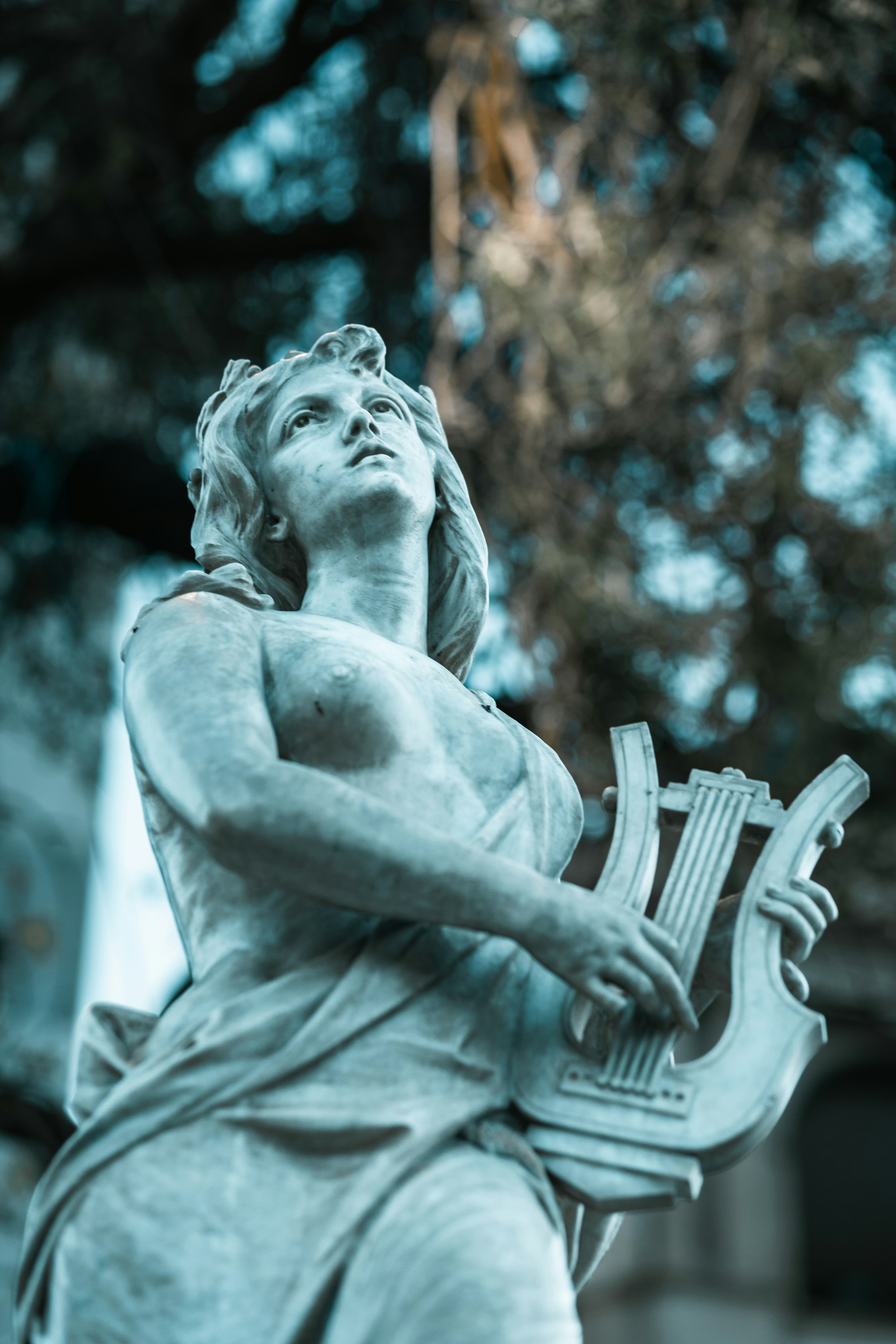 Statue in soft teal glow holds a lyre-like instrument amid a shaded garden