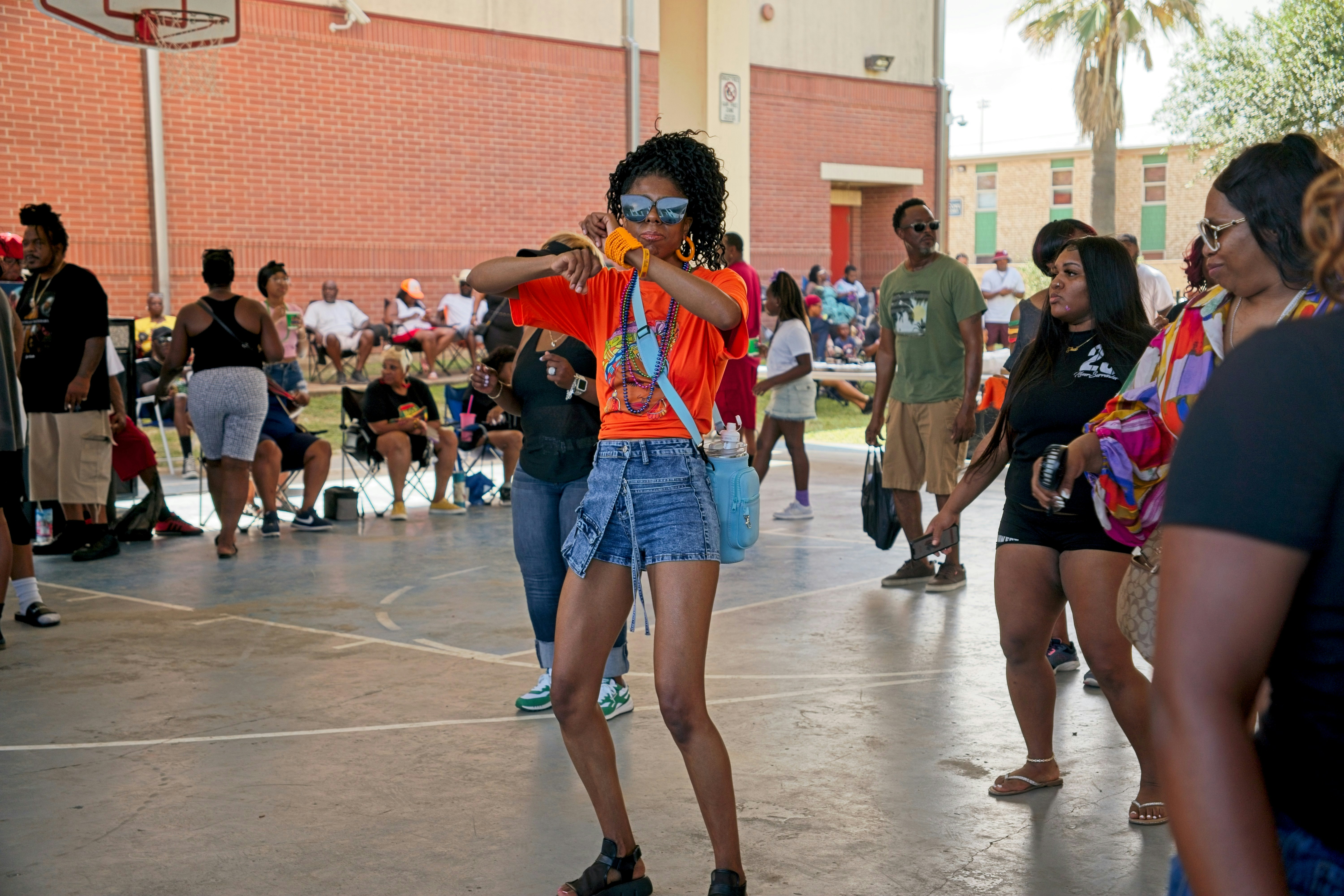 Energetic dancer in a red shirt and denim shorts performs in a busy outdoor gymnasium with onlookers in the background. The scene captures movement and a community event vibe; a logo or branding is not prominently visible.
