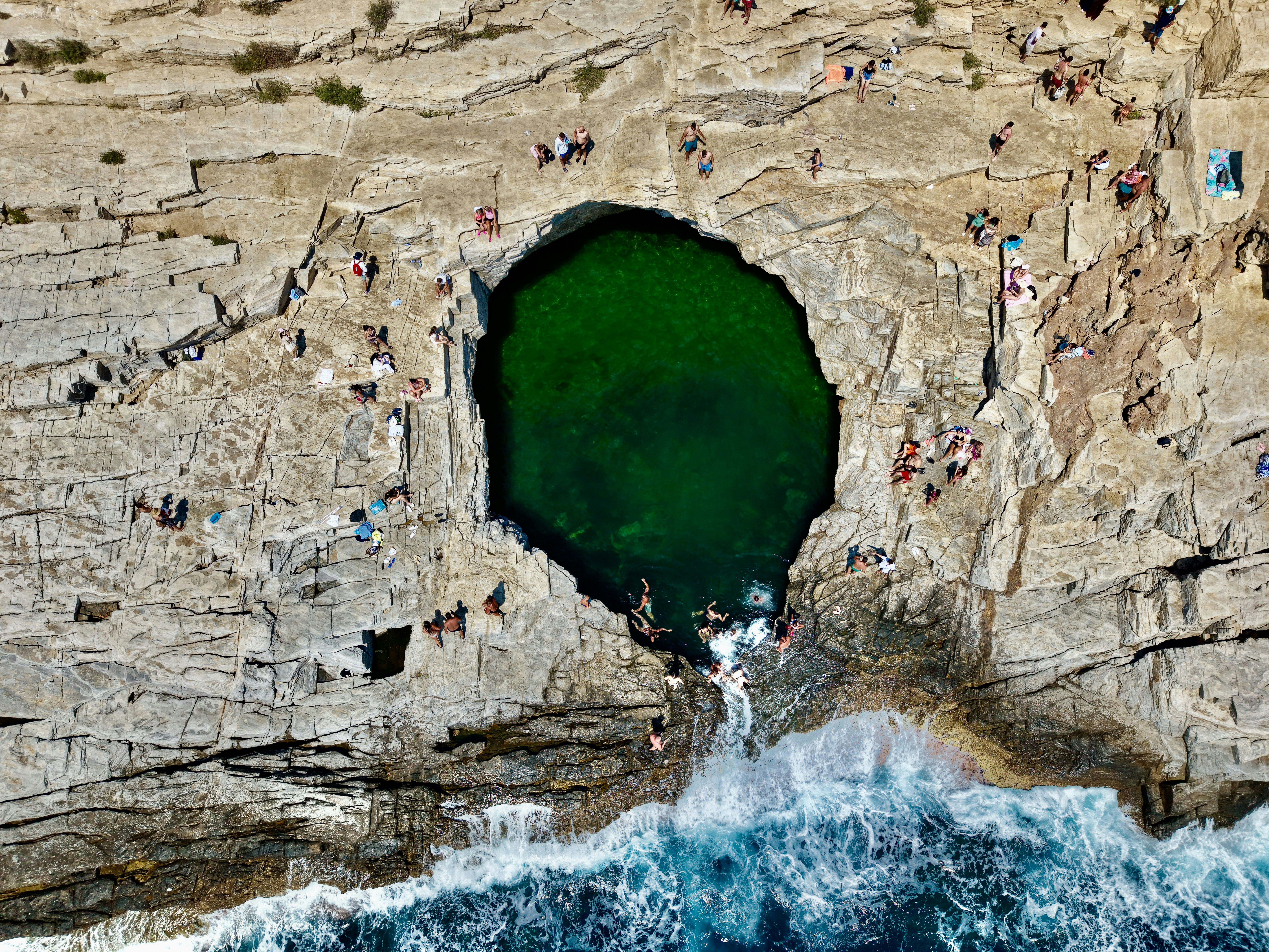 Aerial view reveals a vibrant emerald-green pool set within a circular rock cavity, surrounded by sunlit cliff faces and sunbathers along the rugged shoreline, with turquoise waves washing against the jagged rocks.