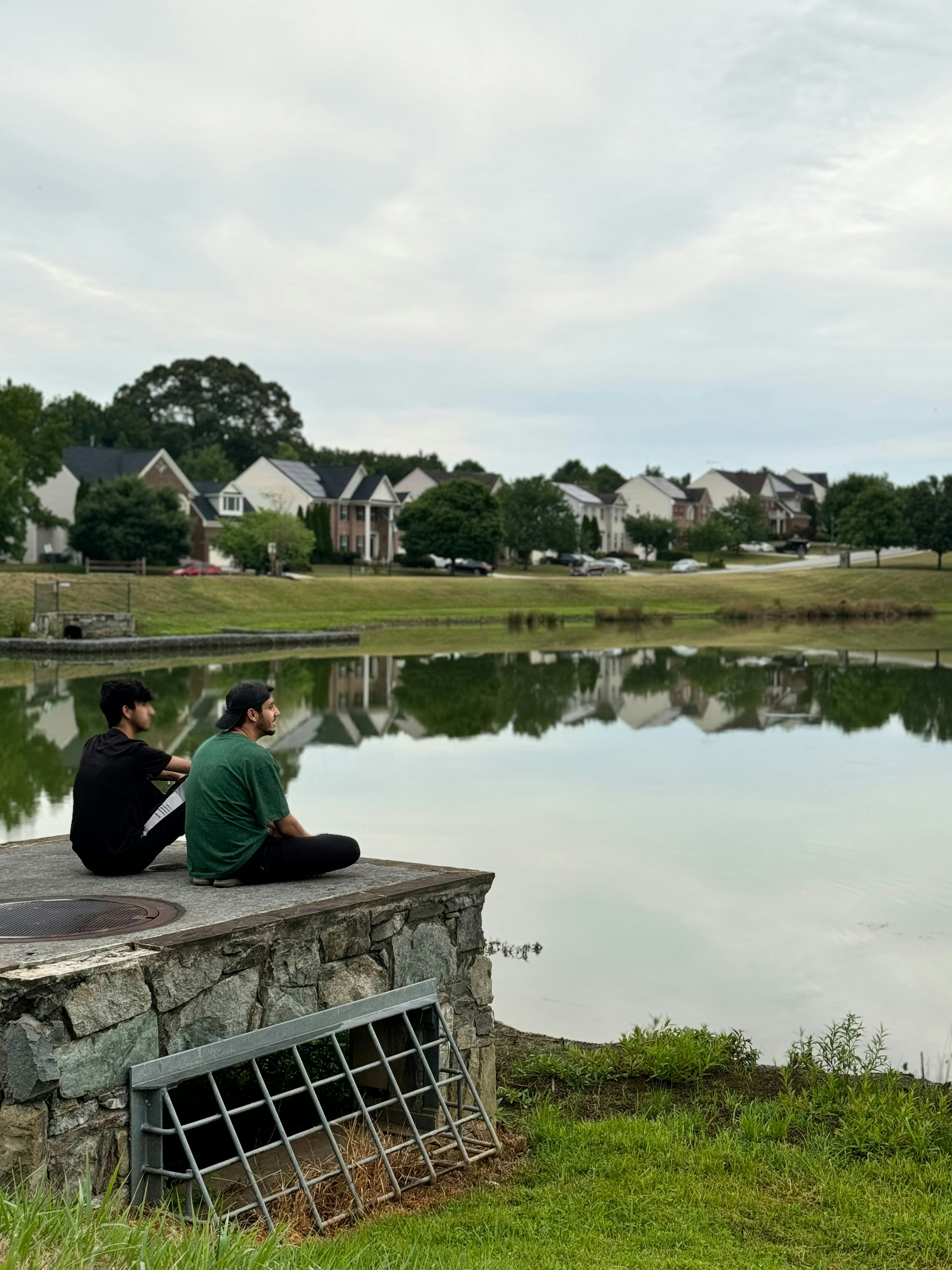 Two men sit on a stone ledge by a calm lake, with a row of suburban houses reflected in the water.