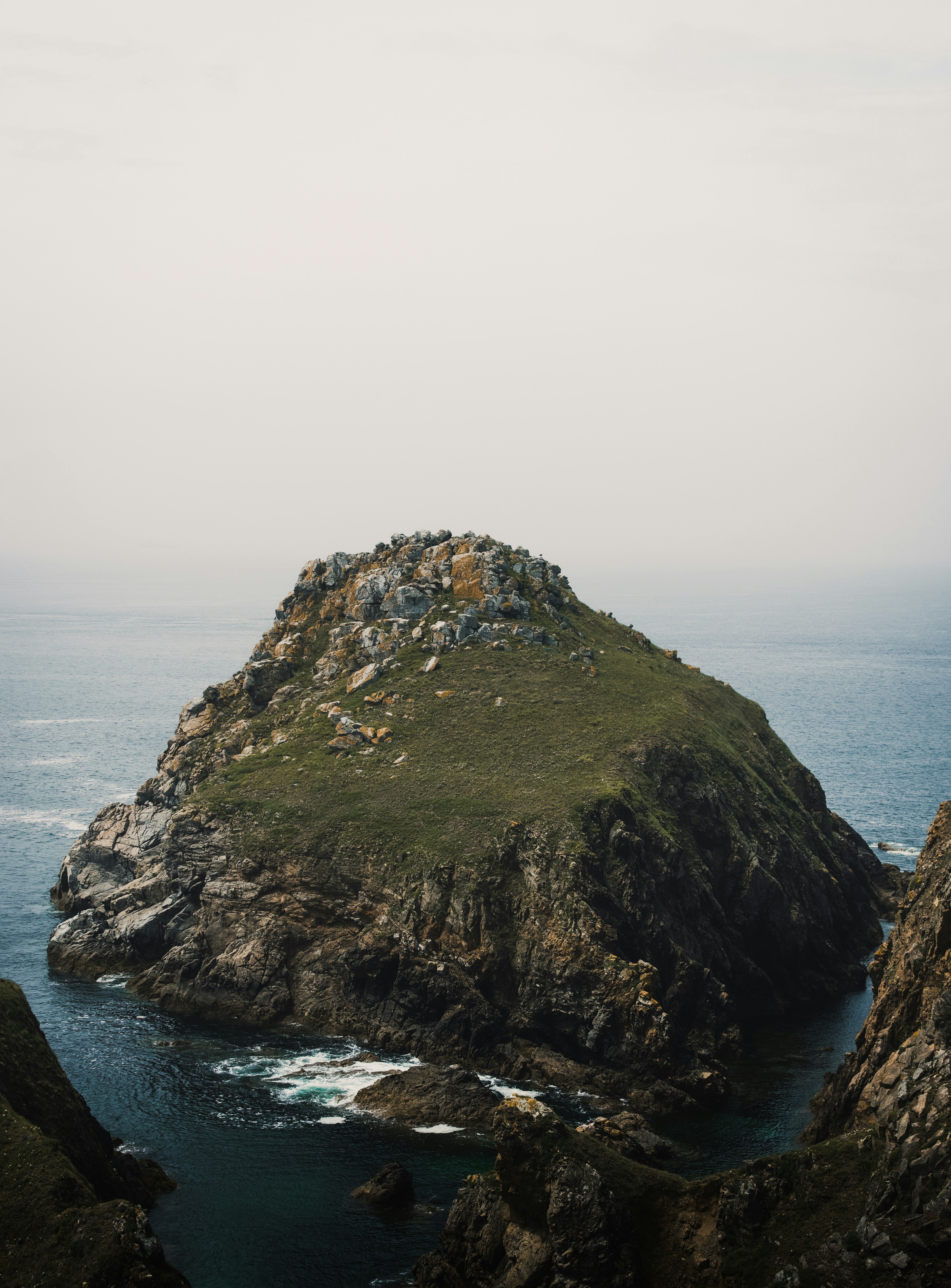 Rocky islet protruding from calm blue water with grassy summit, framed by rugged coastal cliffs.