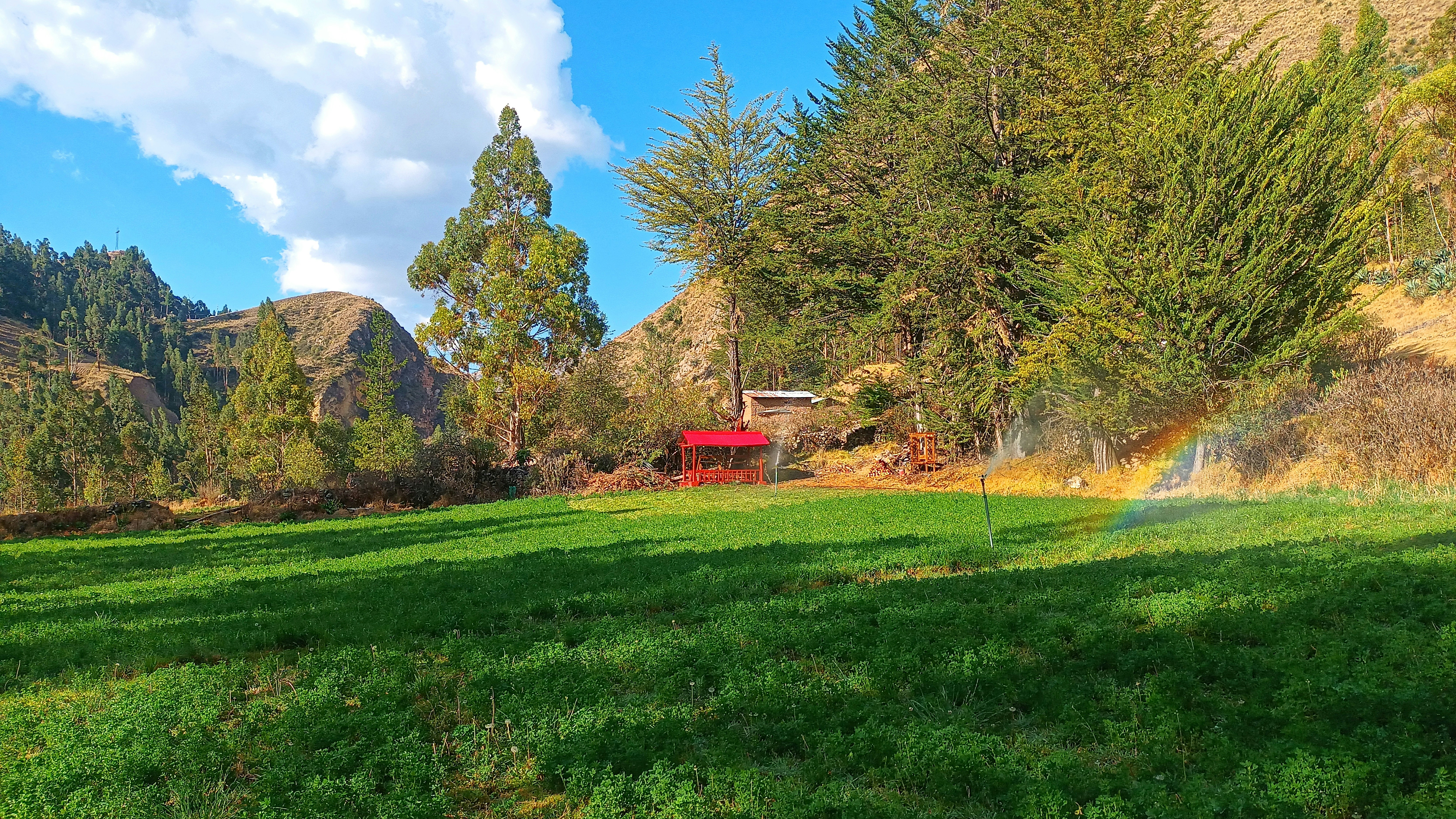Lush green field with a red shelter in the middle ground, framed by trees and distant hills under blue sky with clouds; a faint rainbow arc appears on the right. A bright, natural landscape scene.