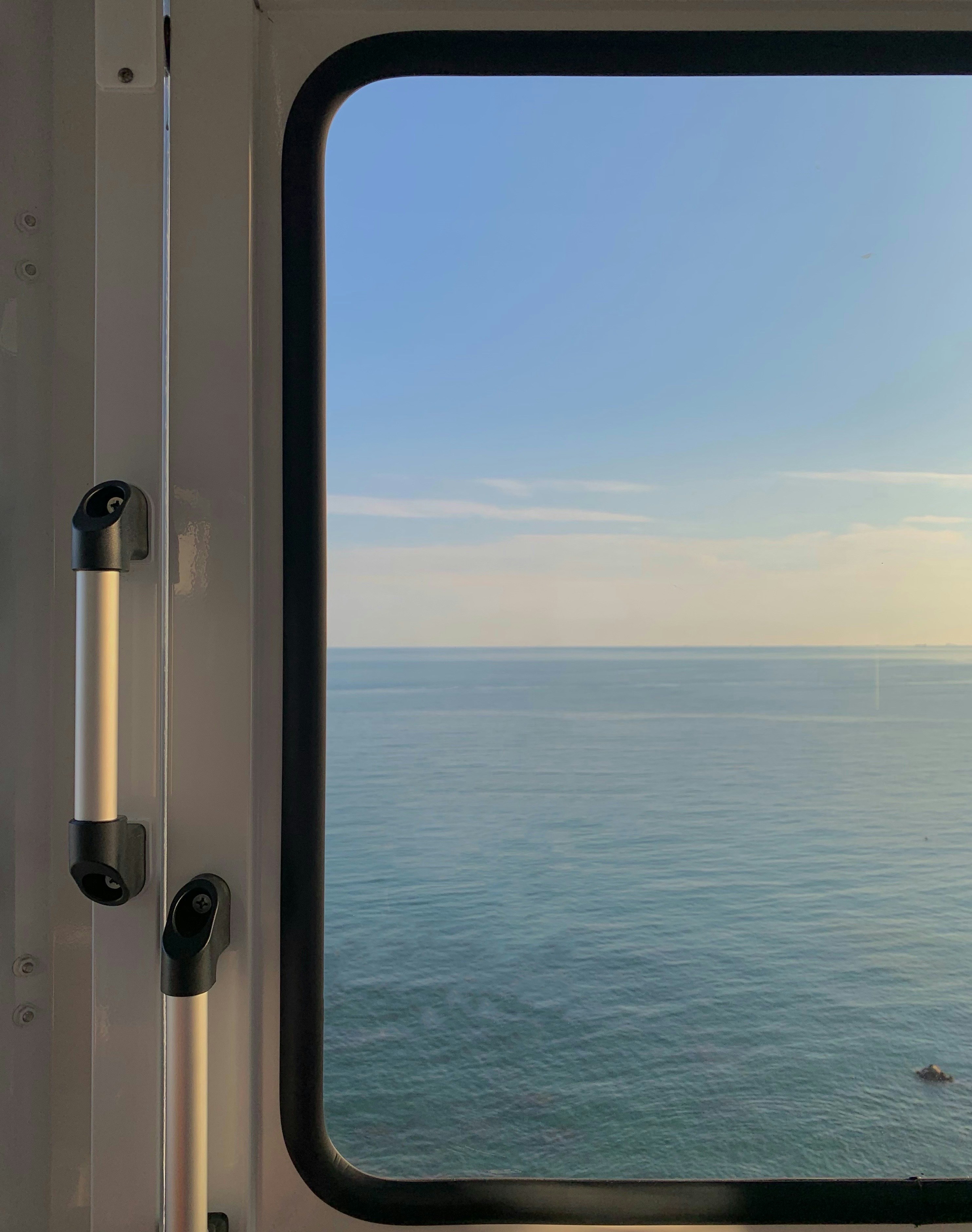 Bright ocean view through a rounded rectangular window with a vertical rail on the left; calm water meets a clear sky at the horizon, suggesting a maritime interior perspective.