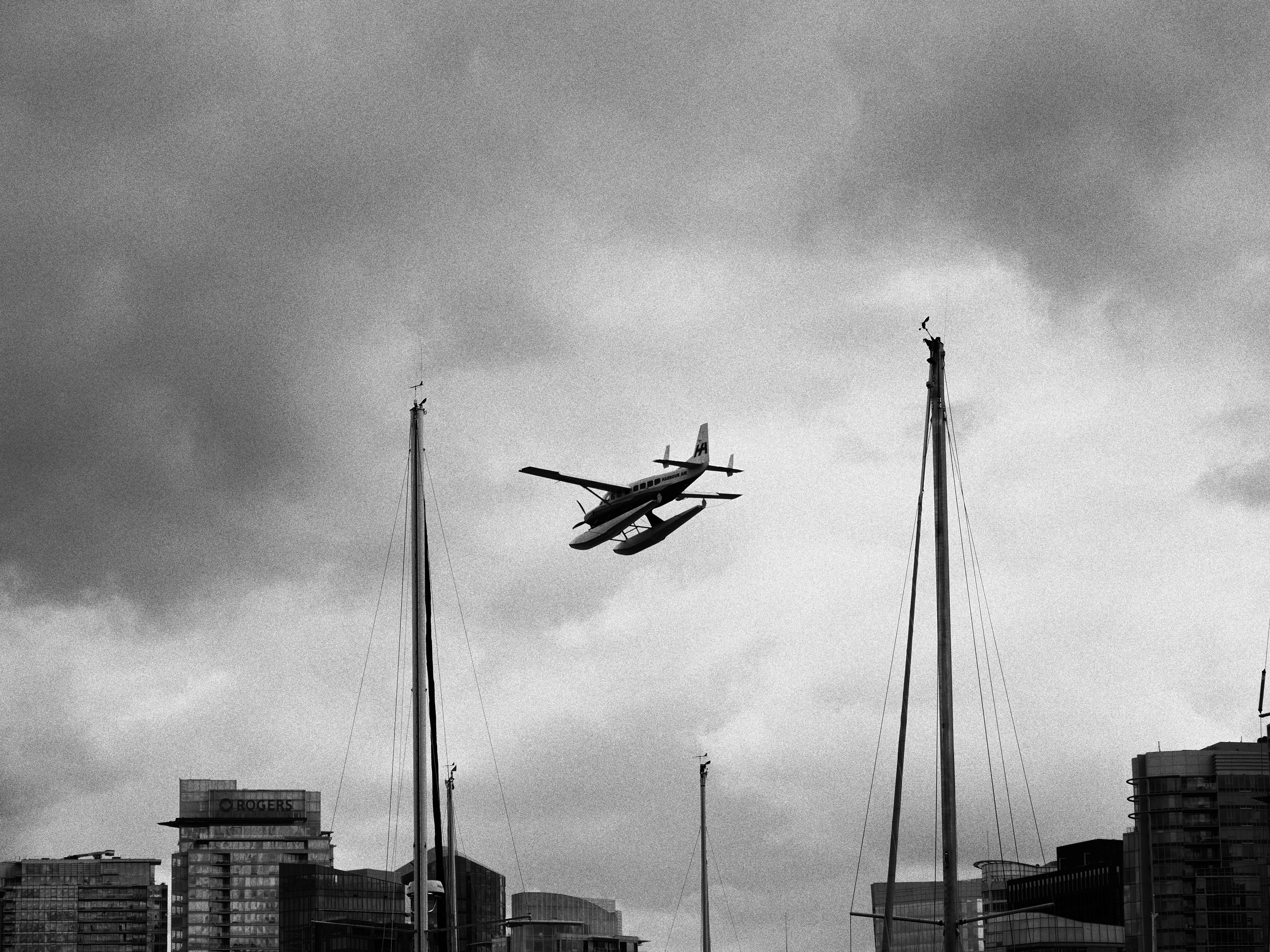 Black-and-white scene of a jetliner performing a low pass or takeoff above a city skyline with tall masts and buildings, capturing motion and contrast in the clouds.