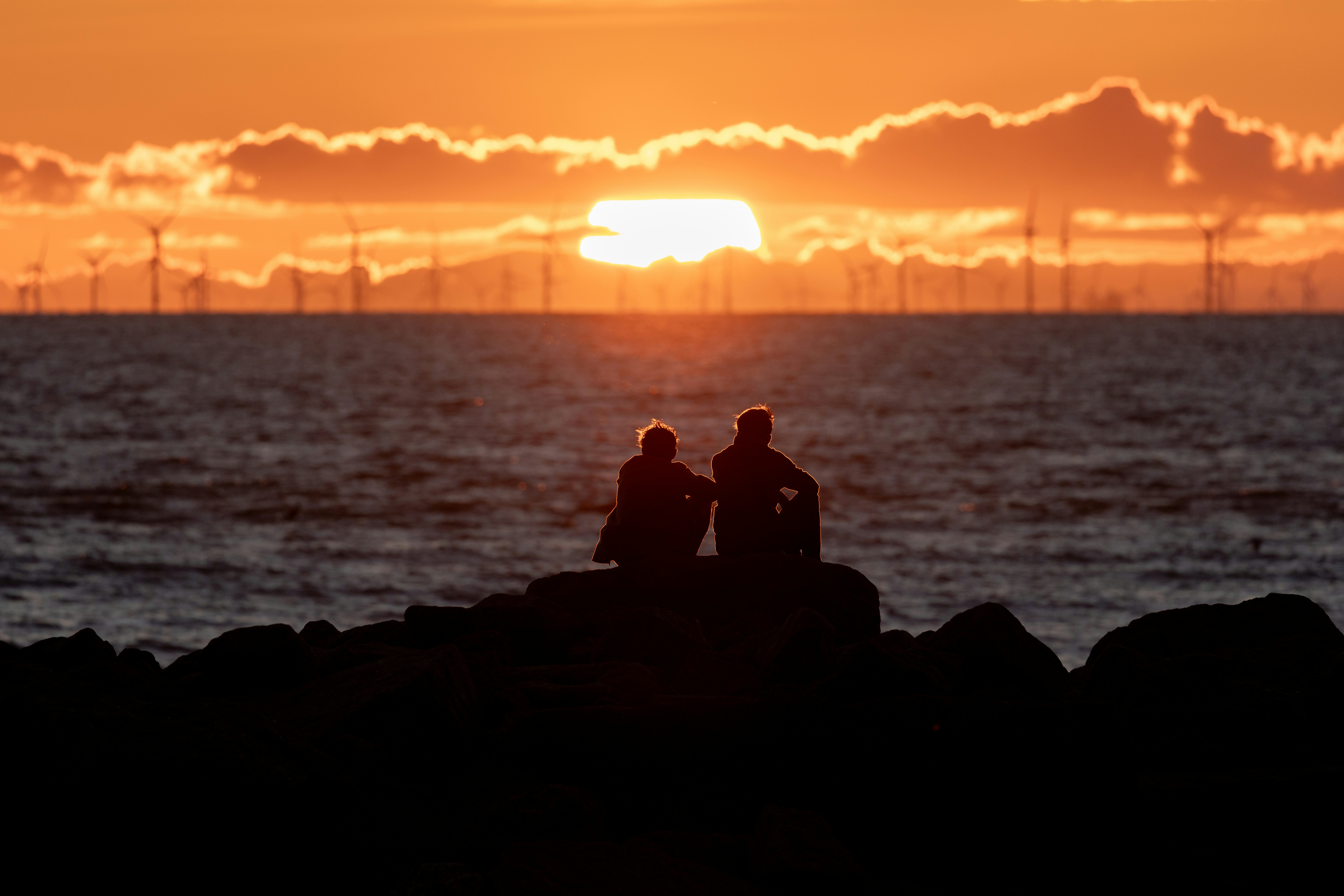 Two silhouetted figures sit on a rocky jetty watching a warm sunset over a calm sea, with distant wind turbines along the horizon.