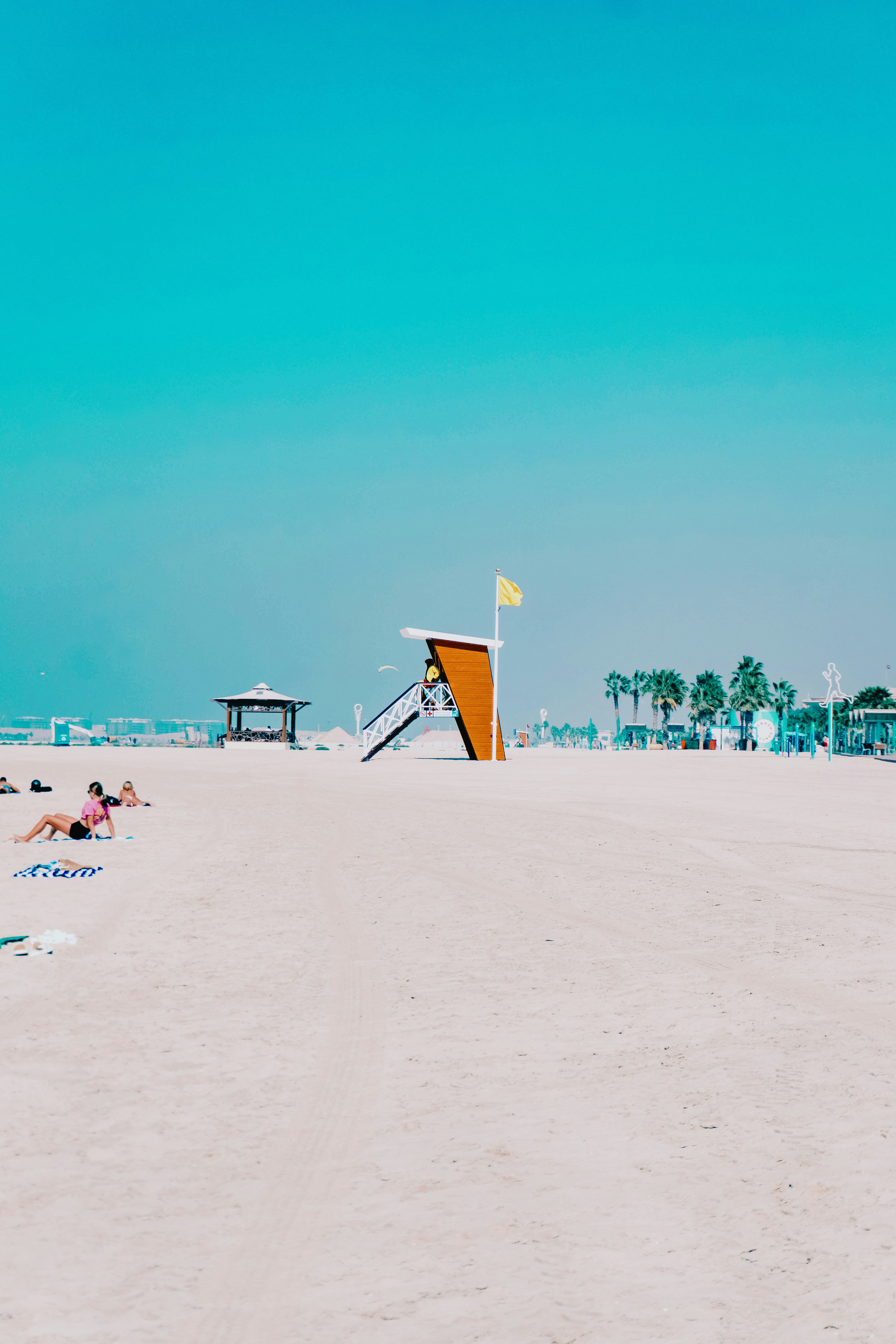 Sunlit beach scene with a solitary lifeguard tower near the horizon and a few sunbathers on the pale sand, under a vivid blue sky.