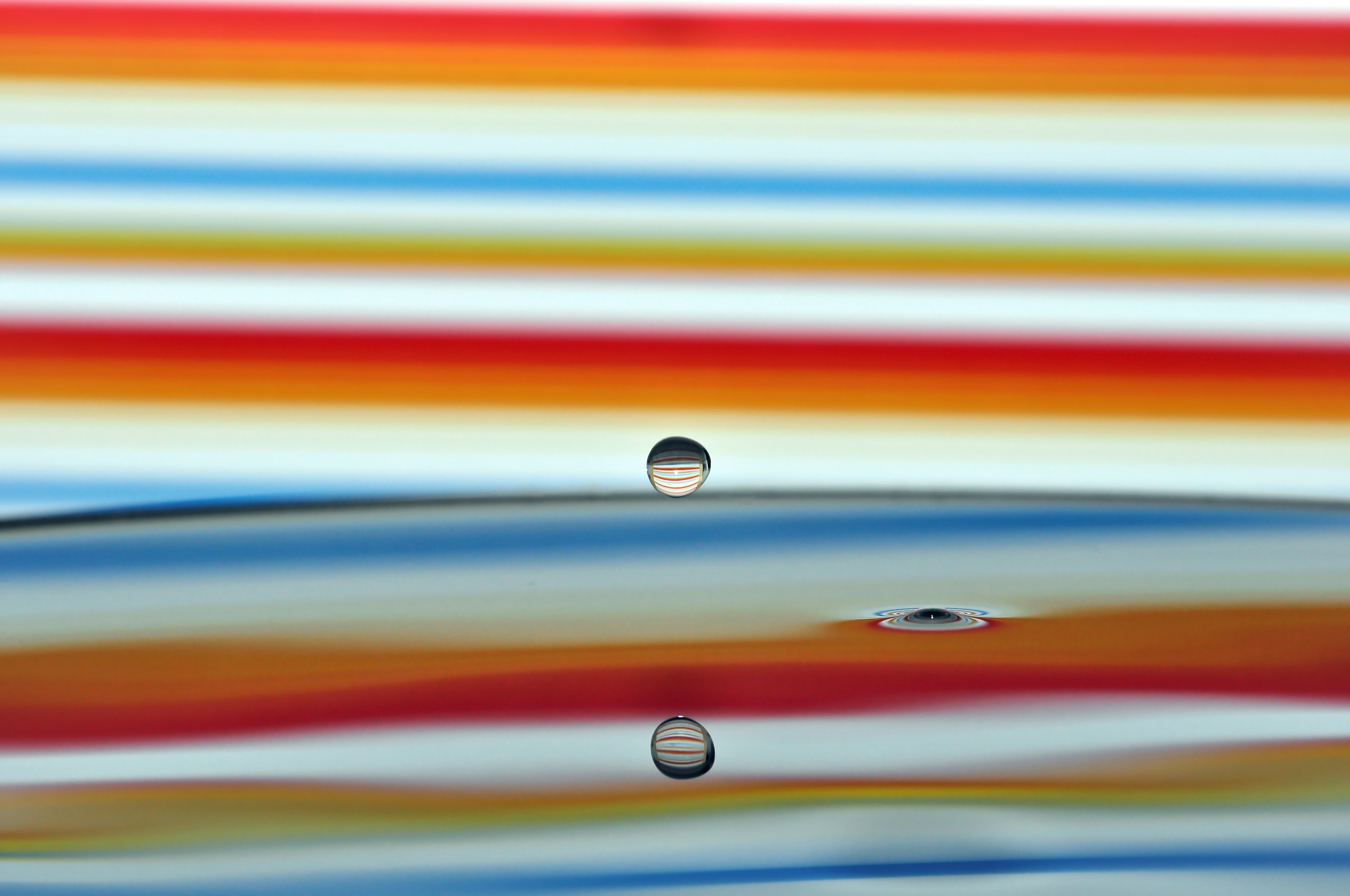 A single water droplet suspended above a rippled surface with colorful horizontal stripes