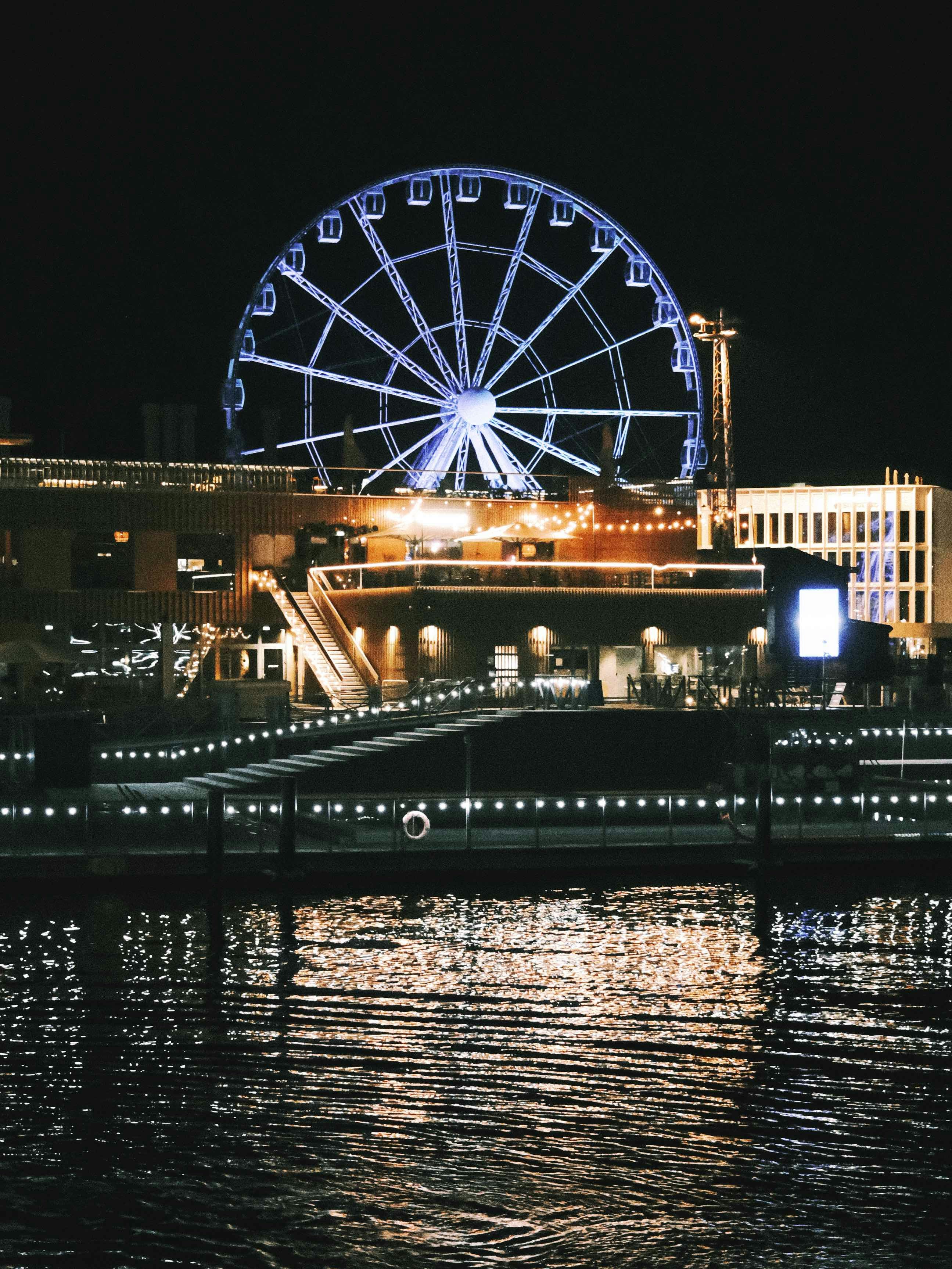 Ferris wheel glows above a lit waterfront complex at night, reflecting on calm water.