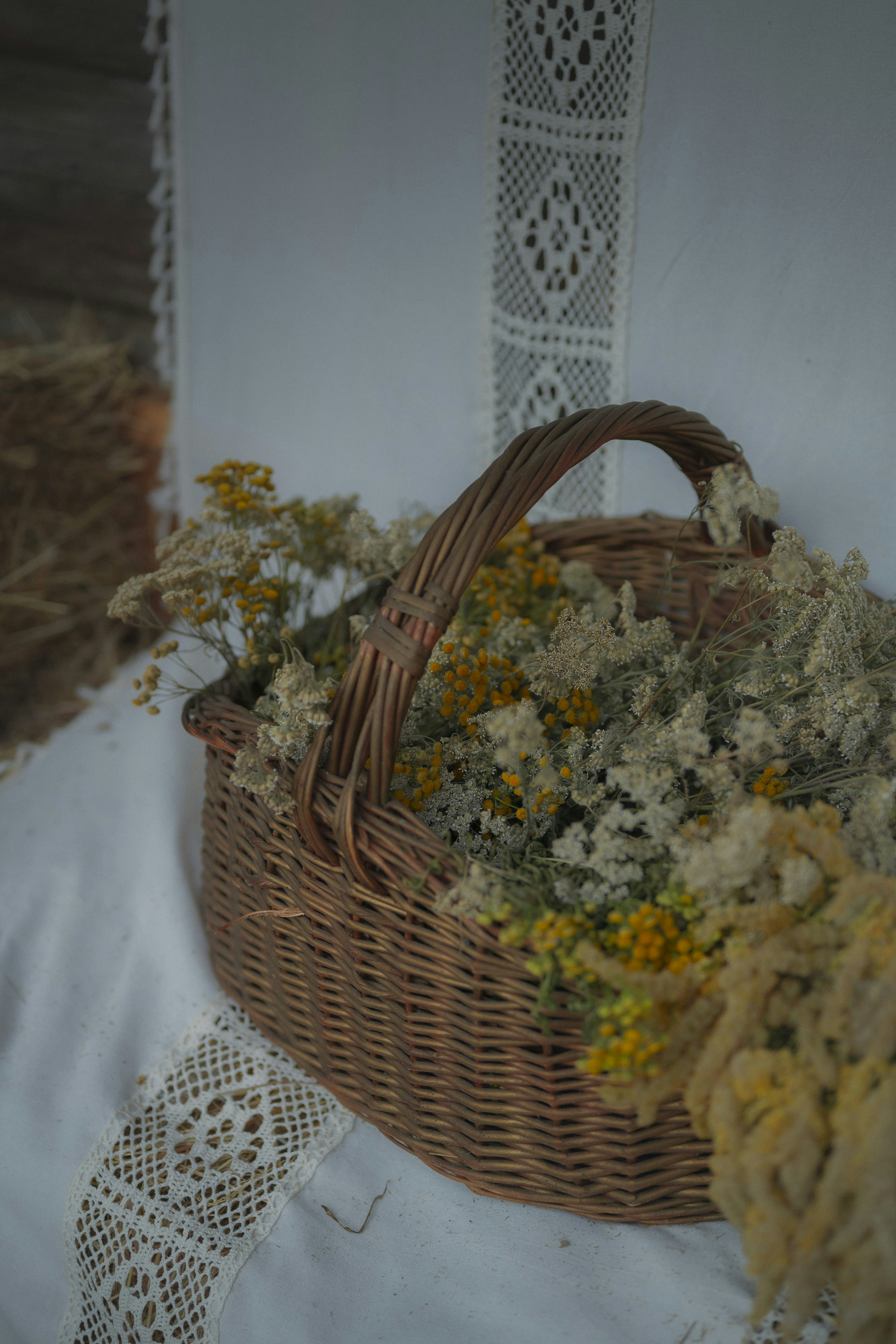 A cozy still-life of a wicker basket filled with wild and dried flowers, set on a white embroidered cloth against a rustic wooden backdrop. Soft, muted tones and natural textures evoke farmhouse charm, cottagecore warmth, and the quiet beauty of a seasonal harvest.