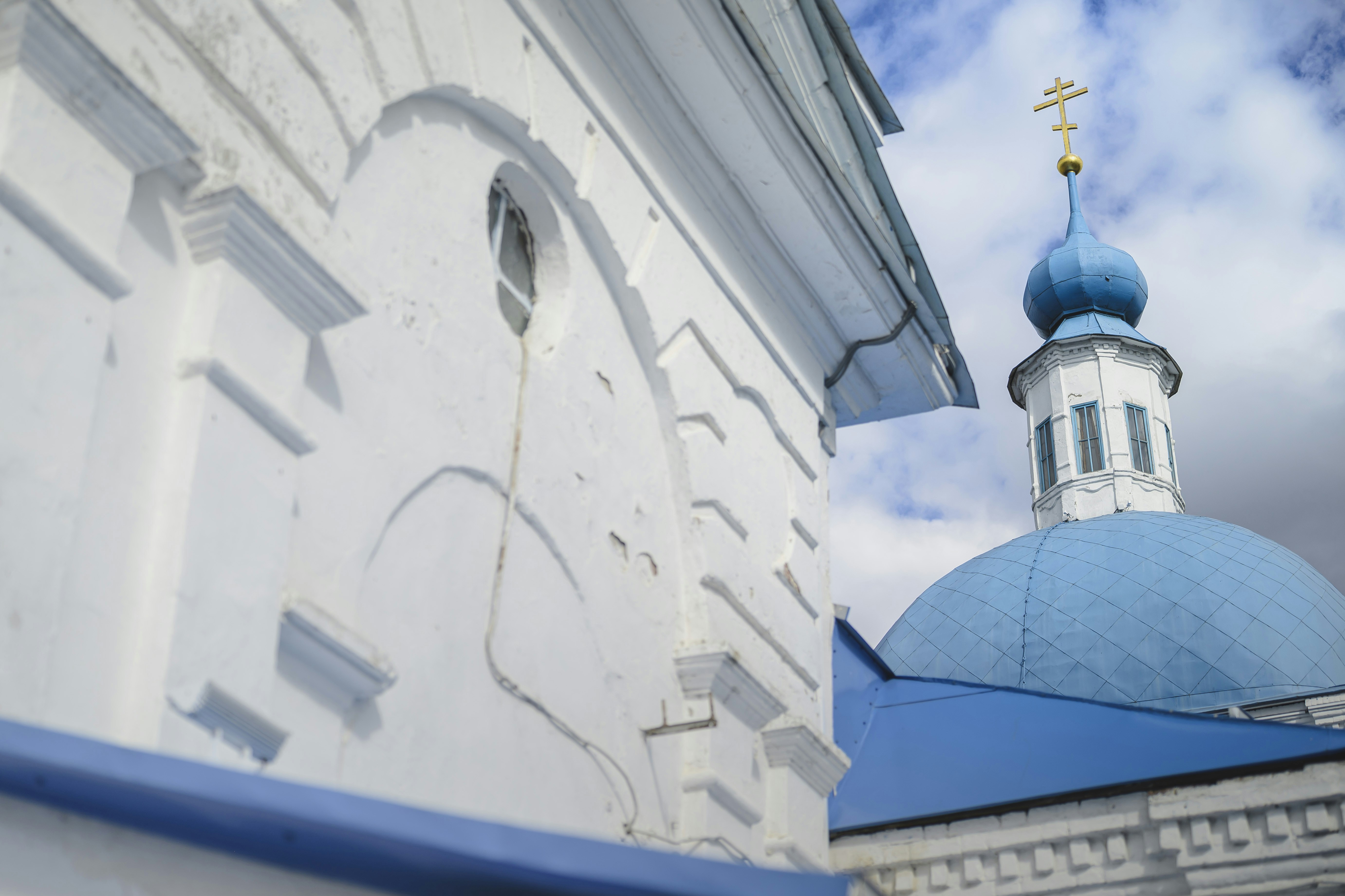 A crisp view of the blue onion dome and golden Orthodox cross of the Church of Elijah the Prophet in Zaraysk, framed against a bright sky. The weathered white façade, delicate cornices and soft cloud backdrop highlight the church’s timeless calm and architectural grace — perfect for travel, architecture, and cultural projects.