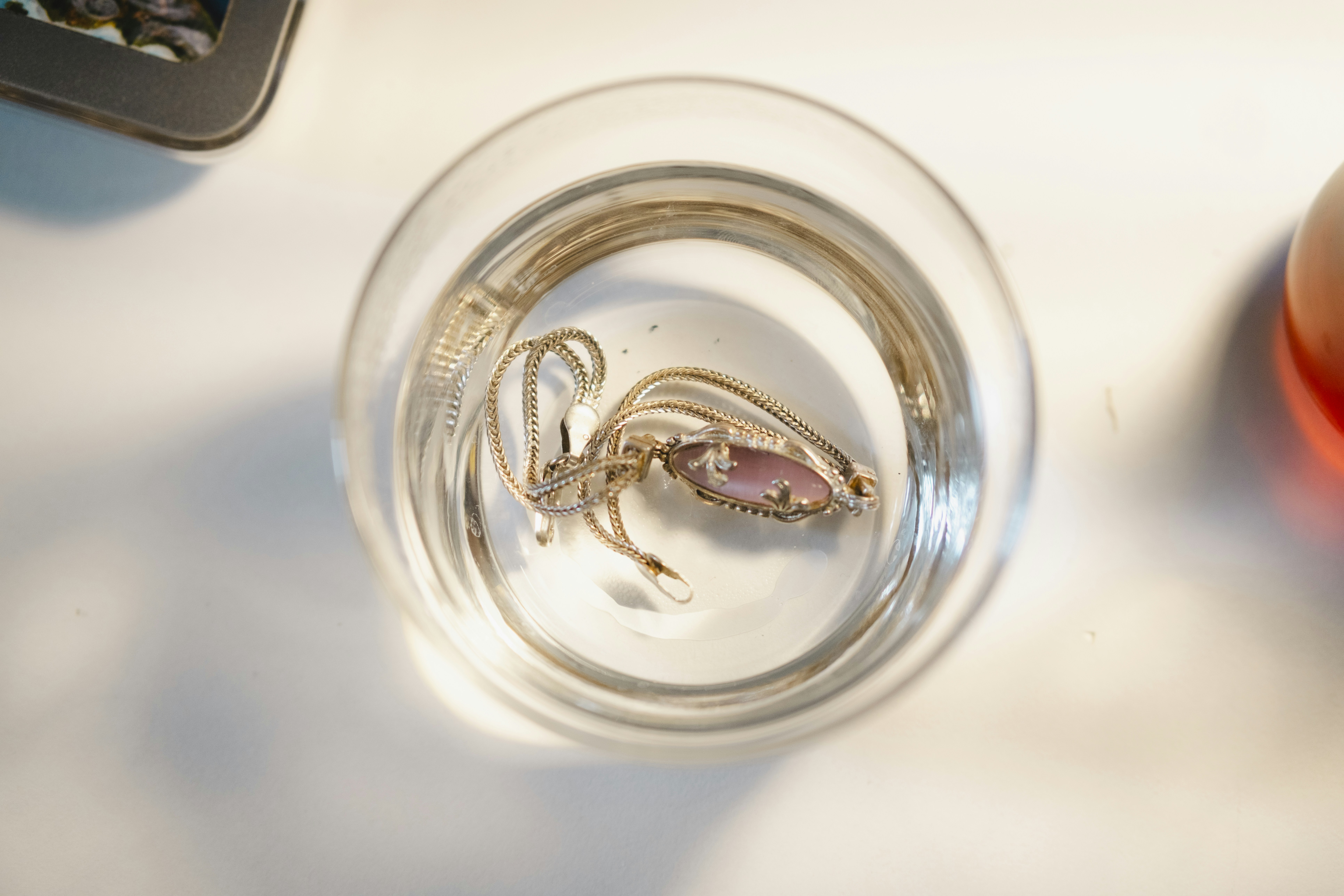 A delicate pendant necklace rests in a glass of water on a tabletop, with soft light and a shallow depth of field. The scene emphasizes the jewelry as the central subject while the surrounding objects blur into a warm backdrop.