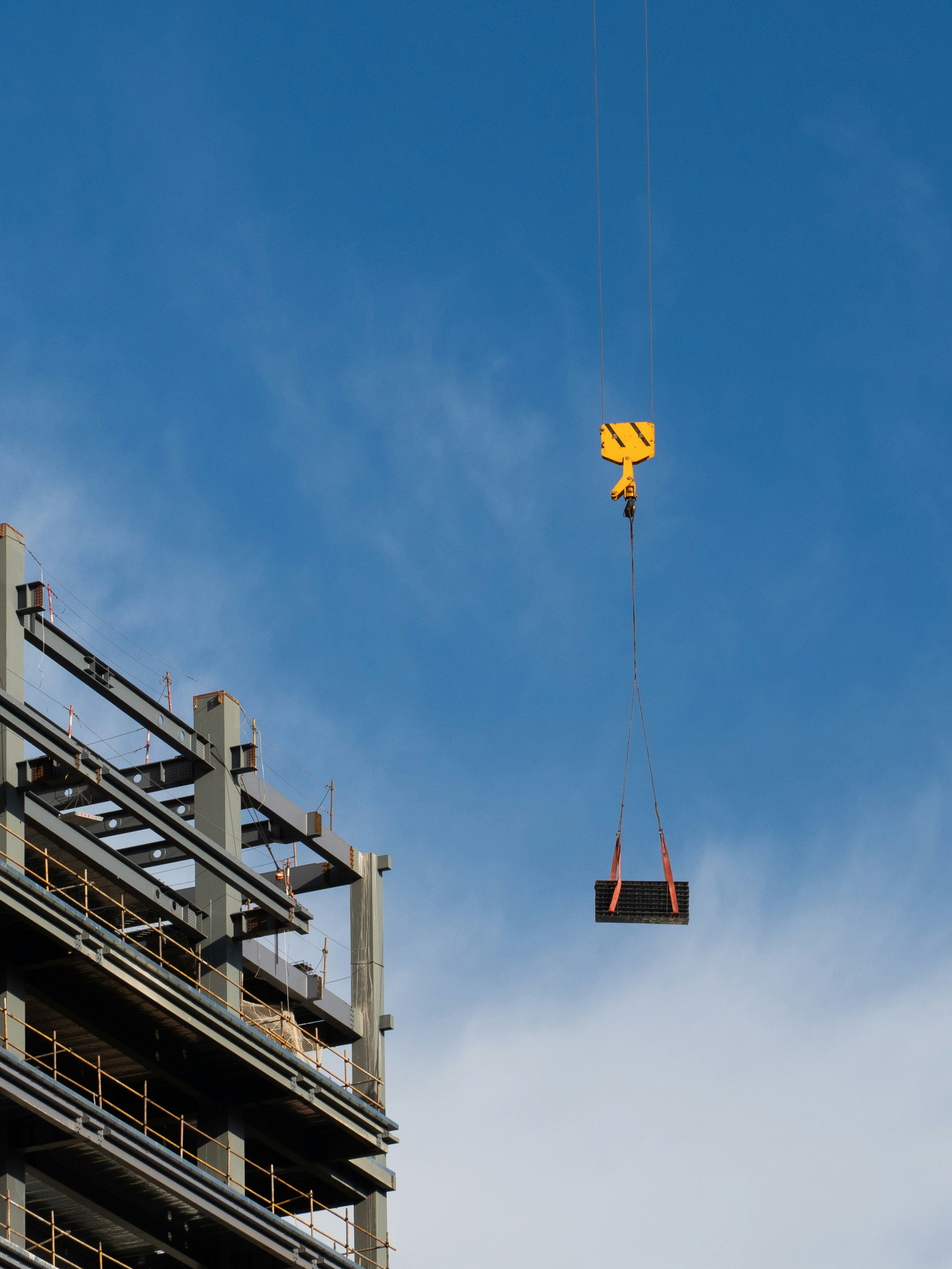 Construction crane hook hoists a load against a bright blue sky, with the skeletal building framework visible at left; minimal clouds, industrial scene. (Logo/pictorial context not indicated.)
