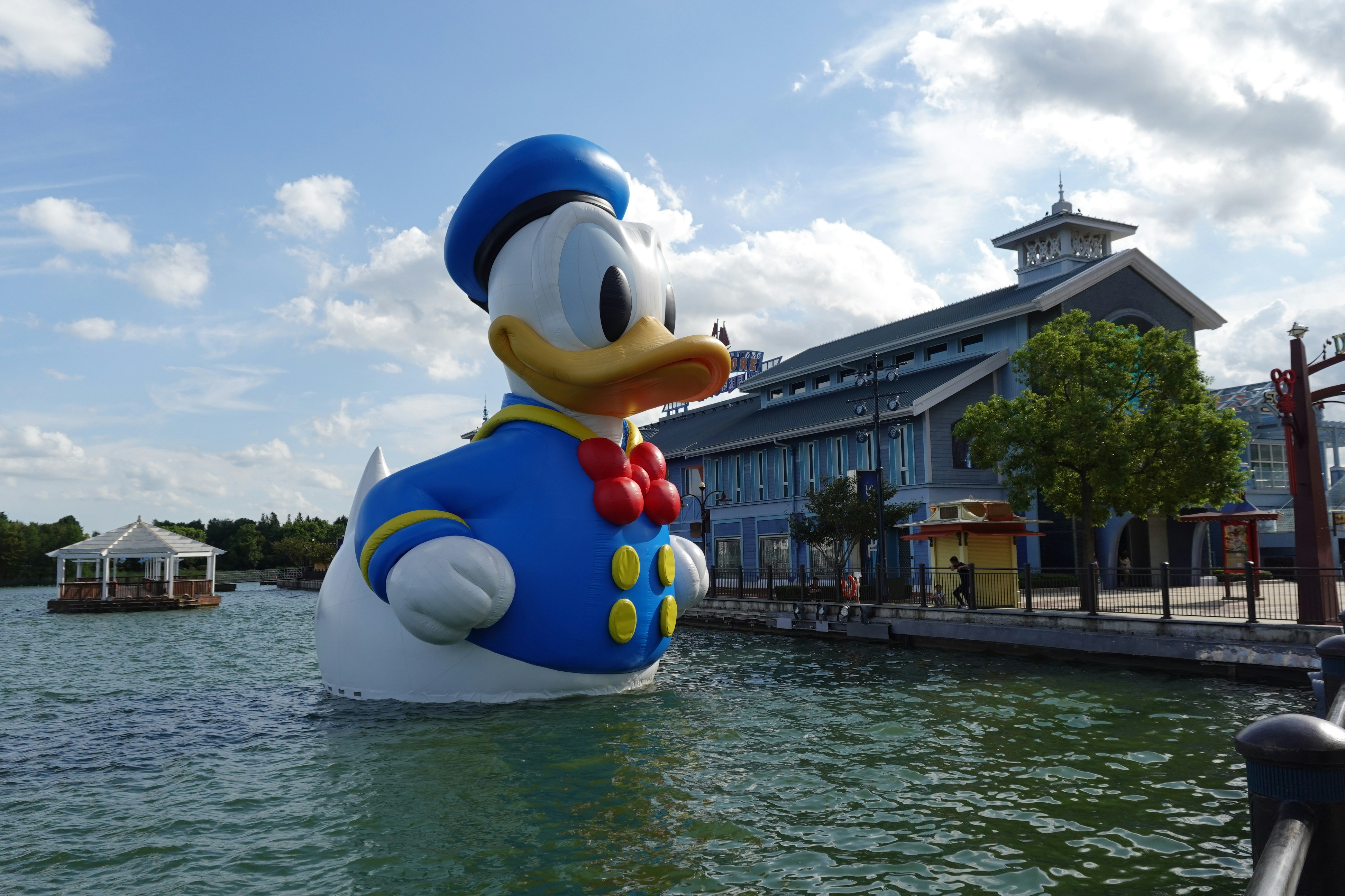 A towering Donald Duck sculpture rises from the water beside a riverside building, creating a playful waterfront centerpiece.