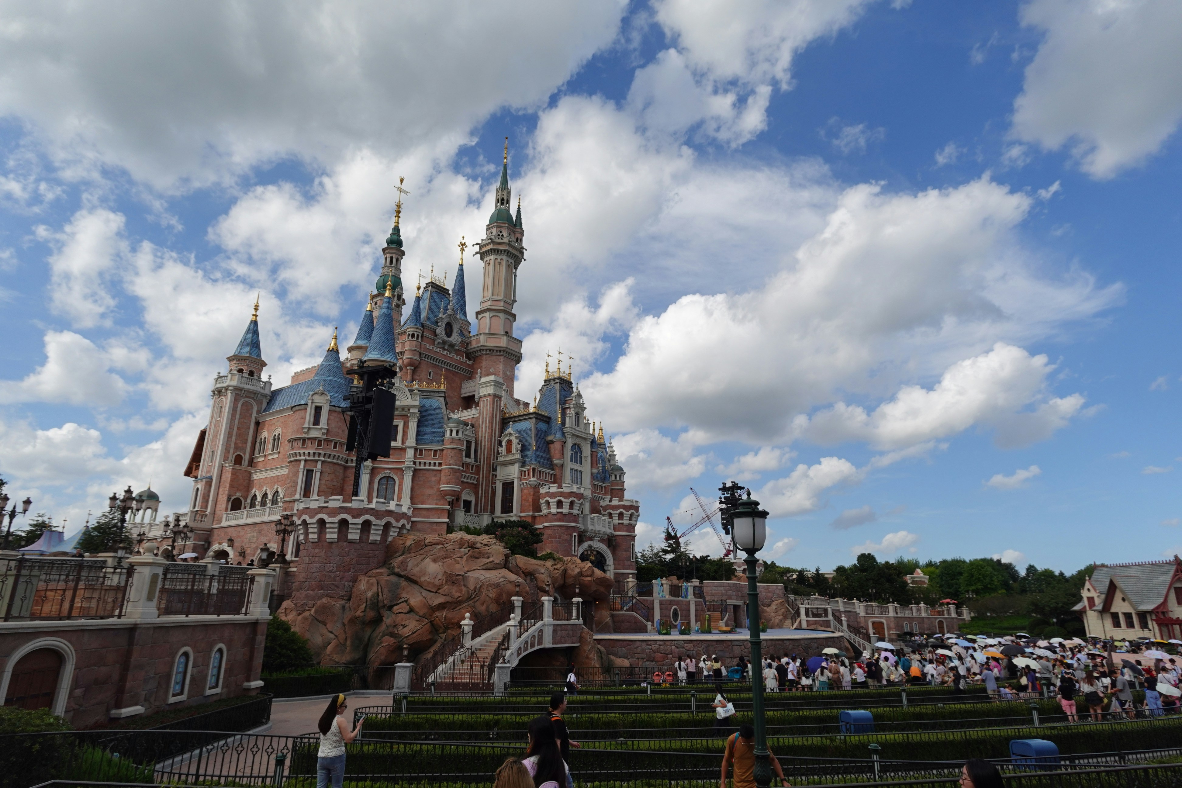 Colorful castle atop a rocky mound under a bright, partly cloudy sky; foreground shows visitors and a well-kept promenade.