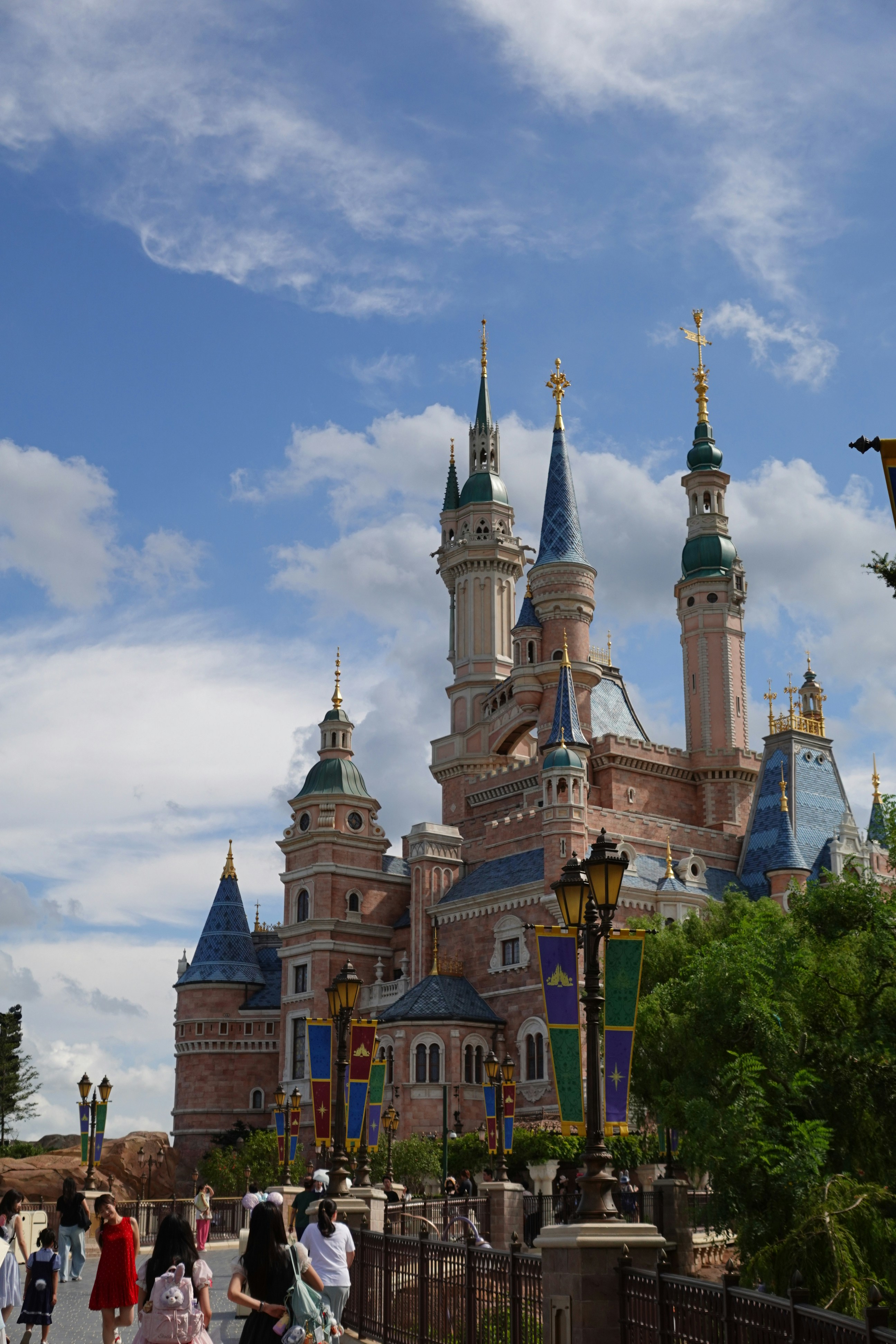 A fairy-tale castle rises against a bright blue sky as guests stroll along a gated promenade; banners line the walkway, and lush greenery frames the scene.
