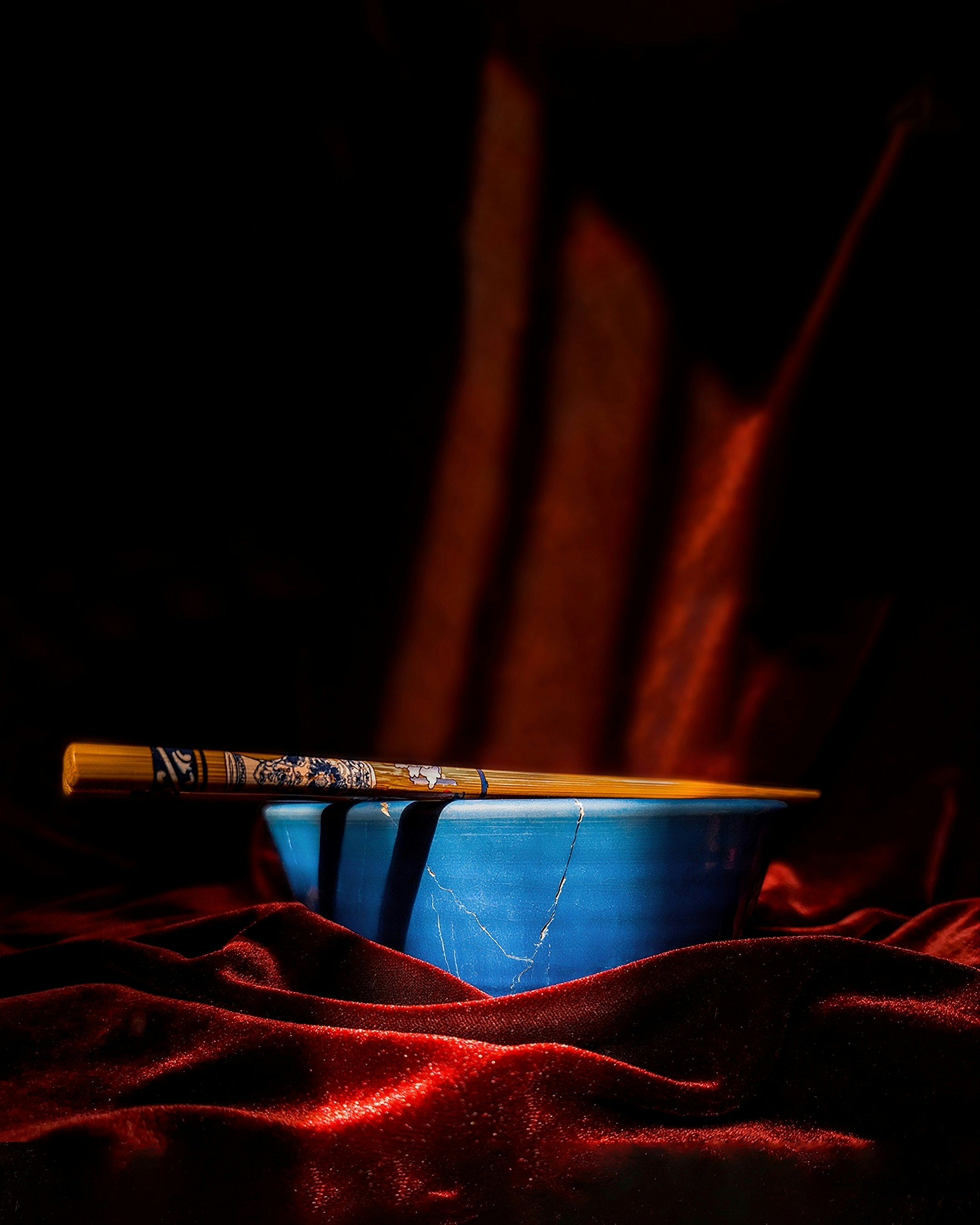 Chopsticks on a blue ceramic bowl A pair of Chinese chopsticks resting on a blue ceramic bowl, surrounded by textured red fabric against a dark background. This dramatic still life combines traditional Chinese cultural elements, ceramic objects, and red lighting as part of a photographic series inspired by the Four Beauties of Ancient China.