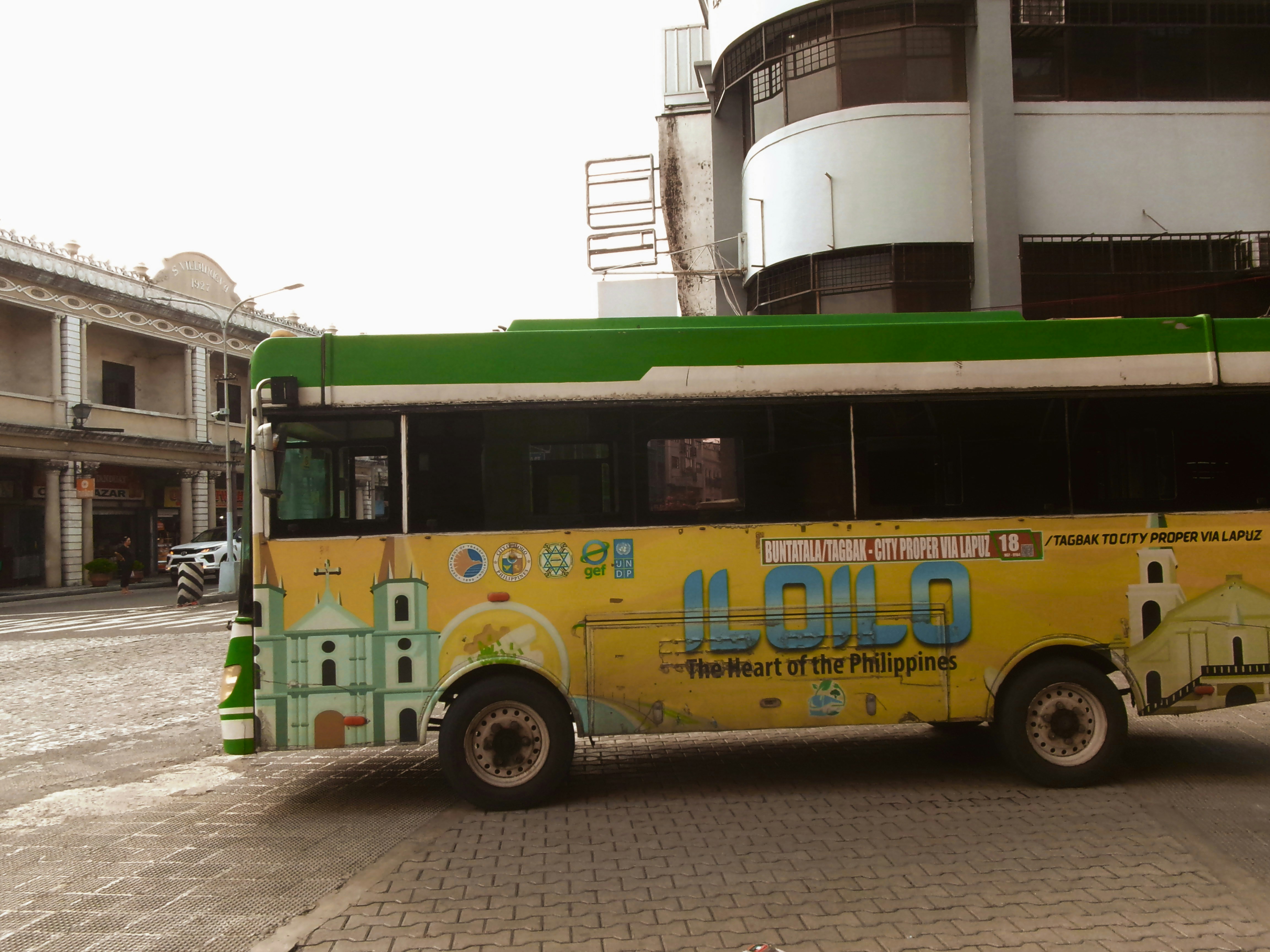 Yellow city bus with green roof sits at a corner, featuring graphic illustrations and text; urban storefronts and a curved white building provide the background.